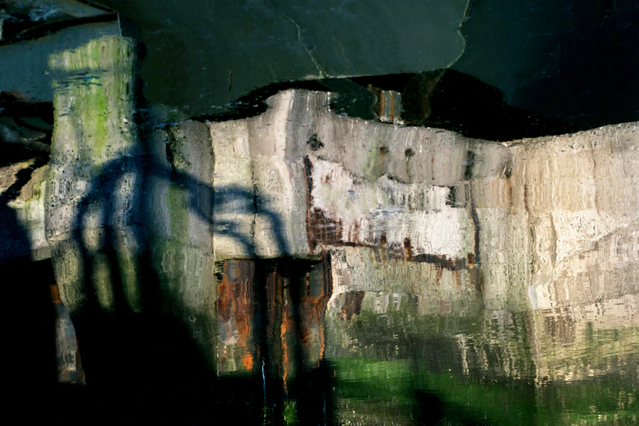 A weathered building facade appears distorted in dark, rippling water with green and blue reflections across the surface.