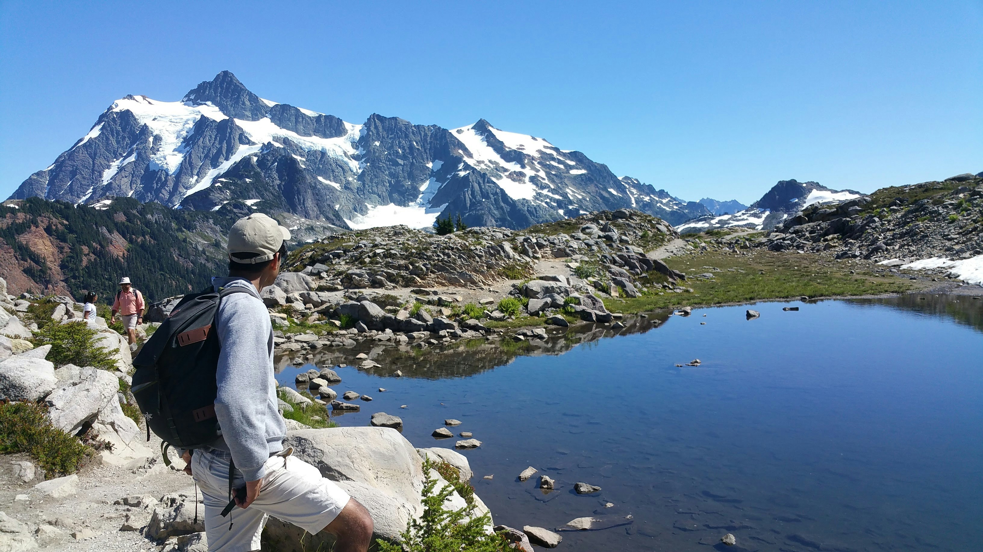 A hiker gazes at the majestic Cascade Mountains, their reflection shimmering in a tranquil alpine pond surrounded by rocky terrain.