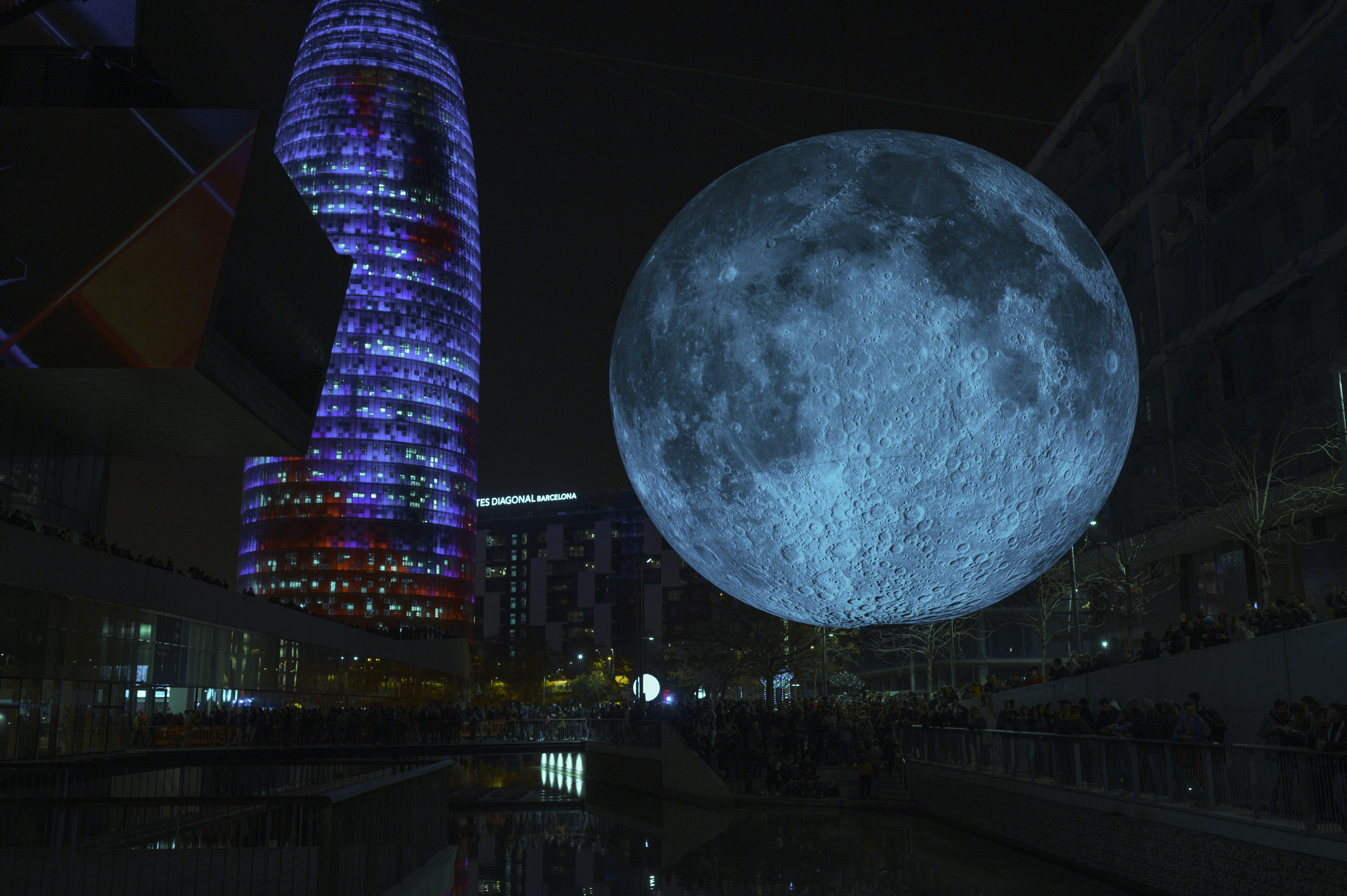 A giant, illuminated moon installation floats beside a vibrant, illuminated skyscraper, creating a stunning contrast against the night sky.