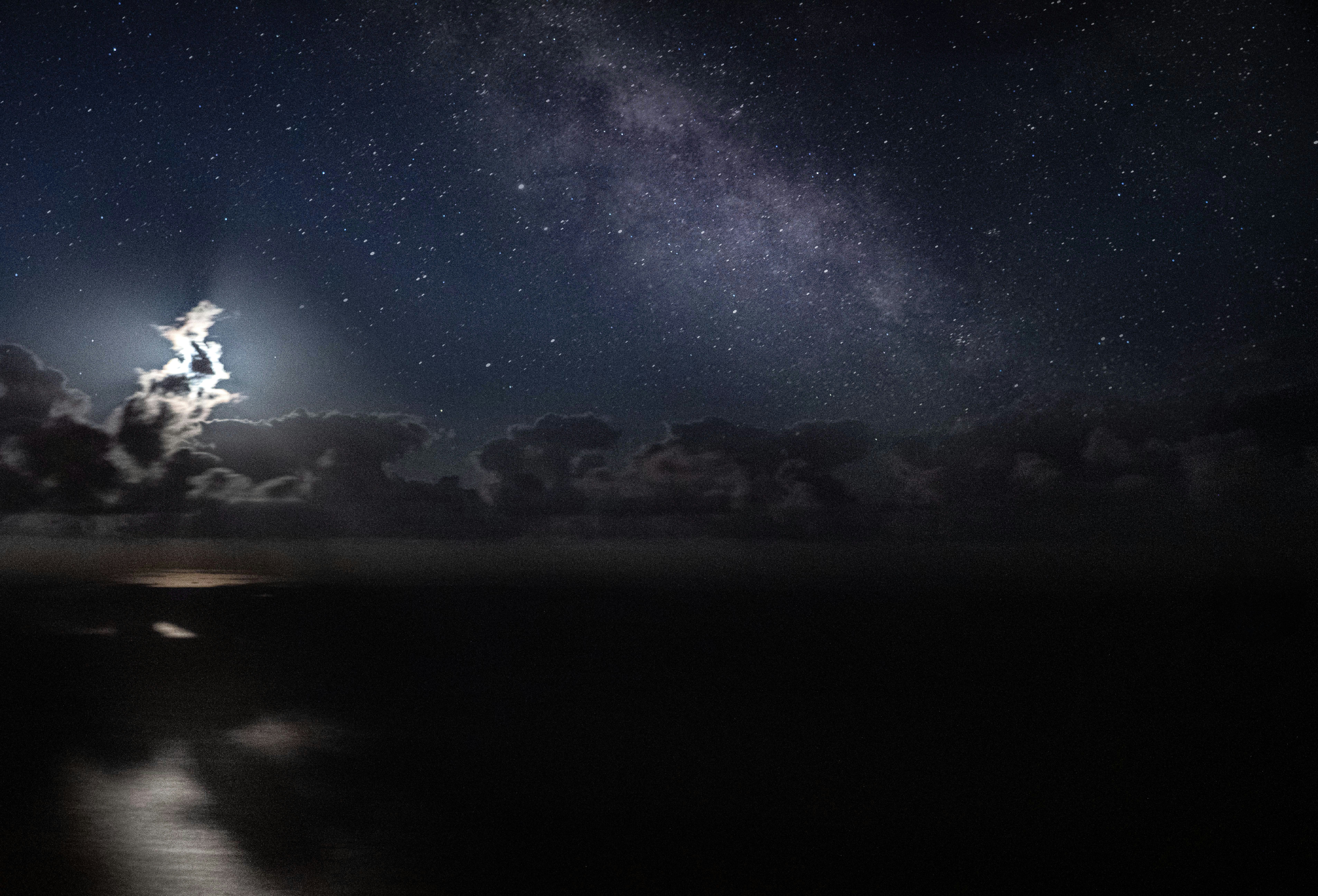 Milky Way arching over a dramatic night sky with illuminated clouds and a reflective ocean surface.
