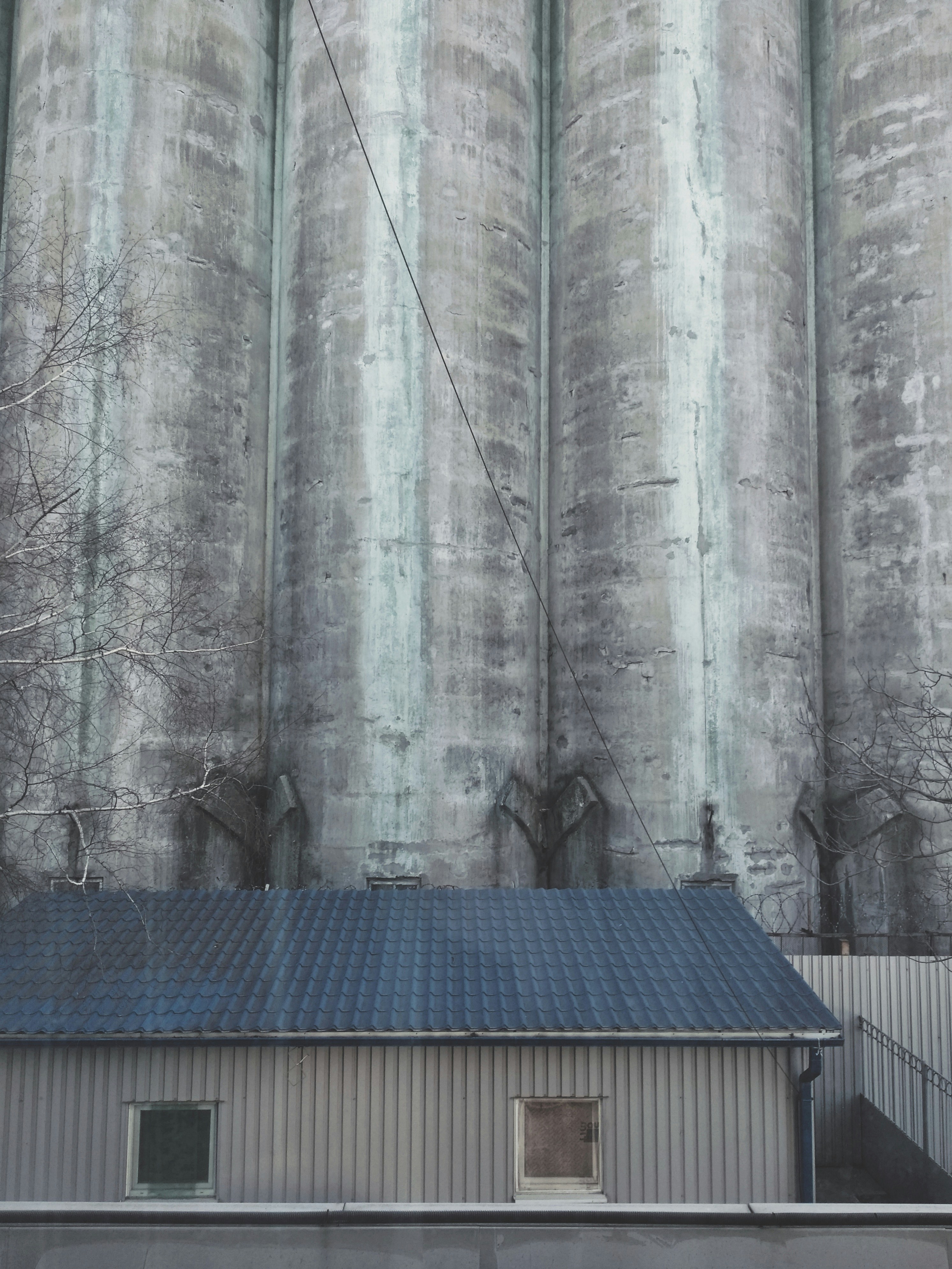 Tall weathered silos rise behind a small corrugated metal shed with a blue roof. A diagonal cable crosses the frame.