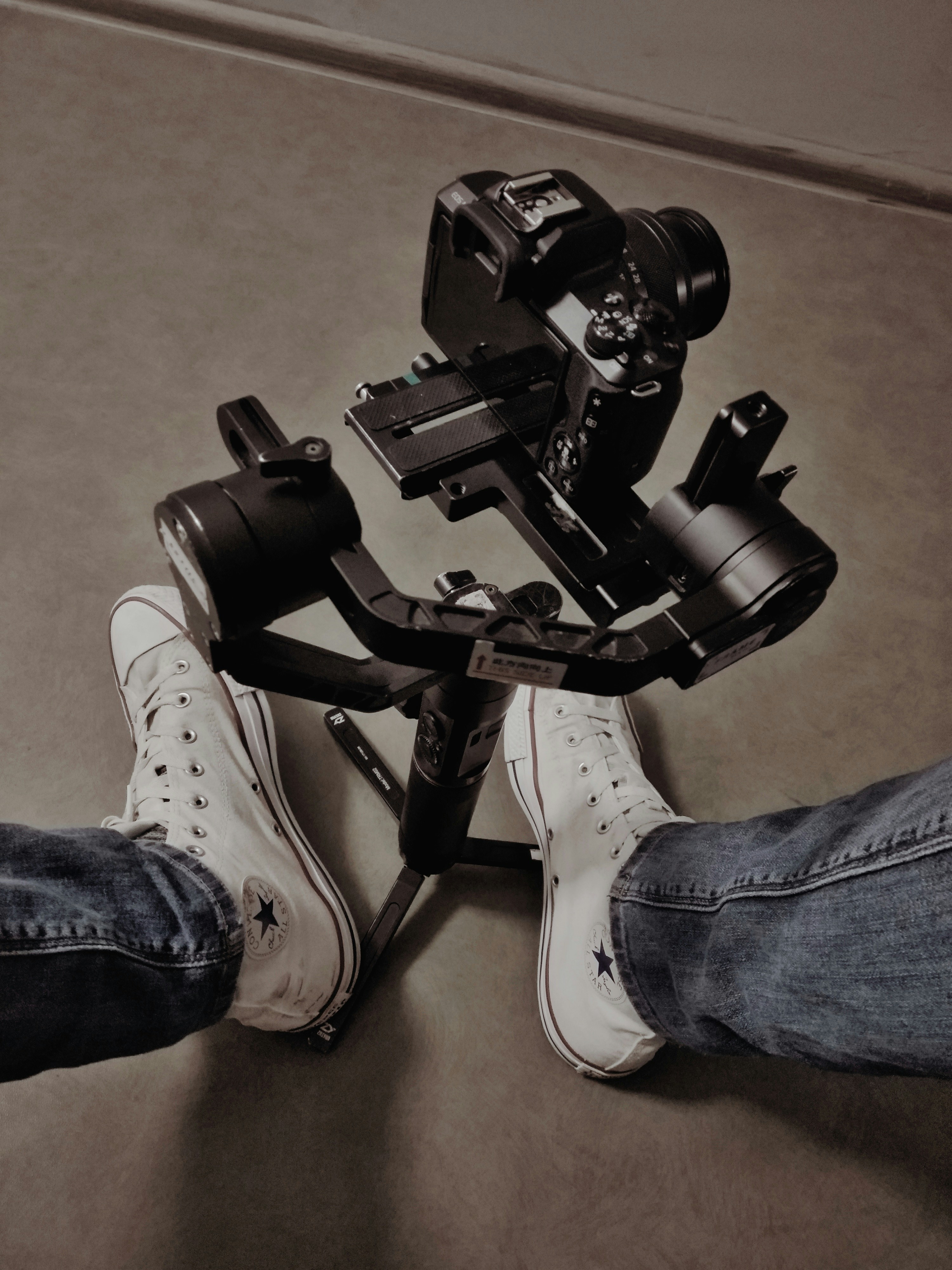 A professional camera stabilizer rests on the floor between the photographer's white sneakers, ready for a shoot.