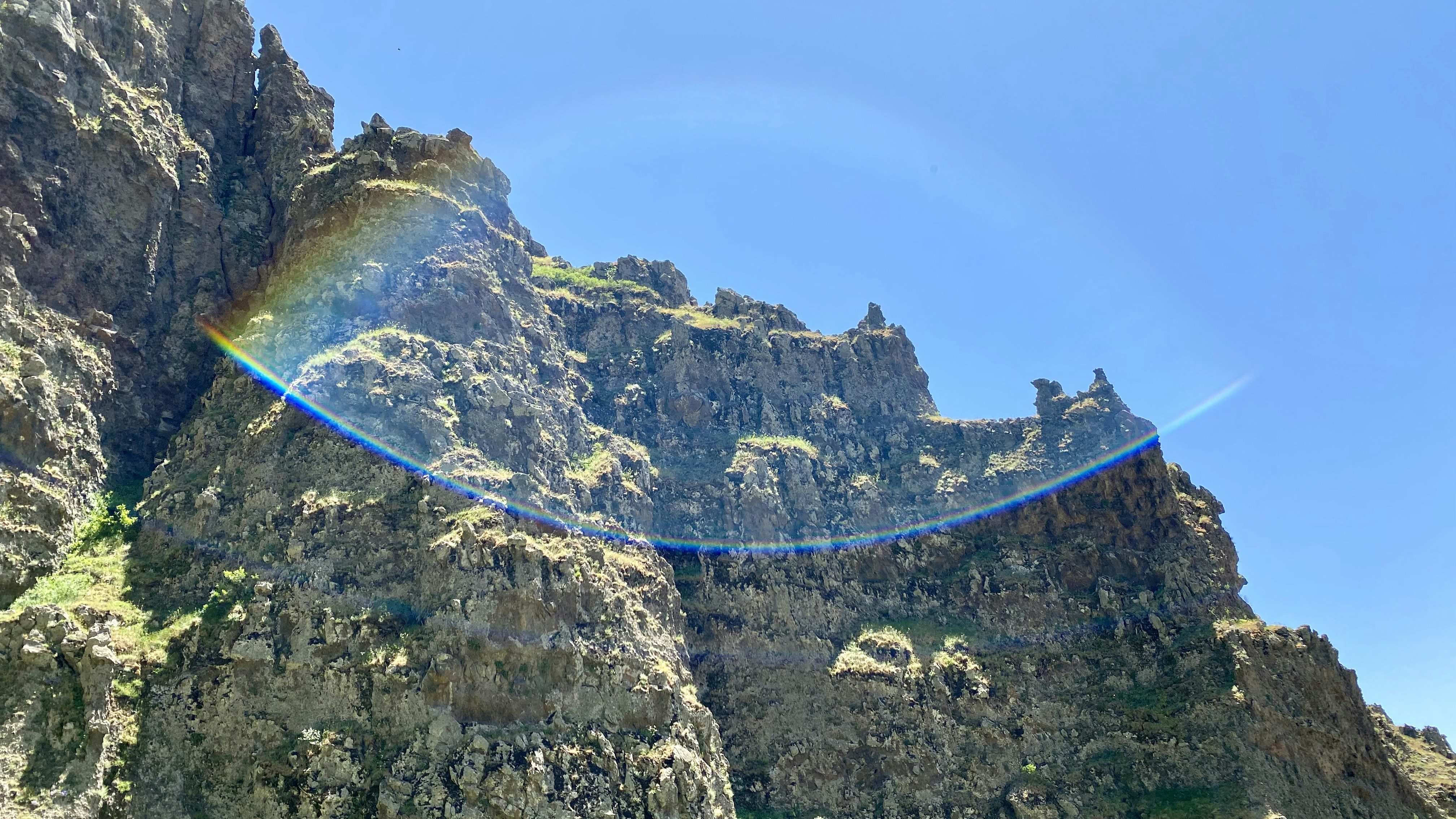 Sunlight creates a rainbow lens flare over rocky mountain cliffs under a clear blue sky.