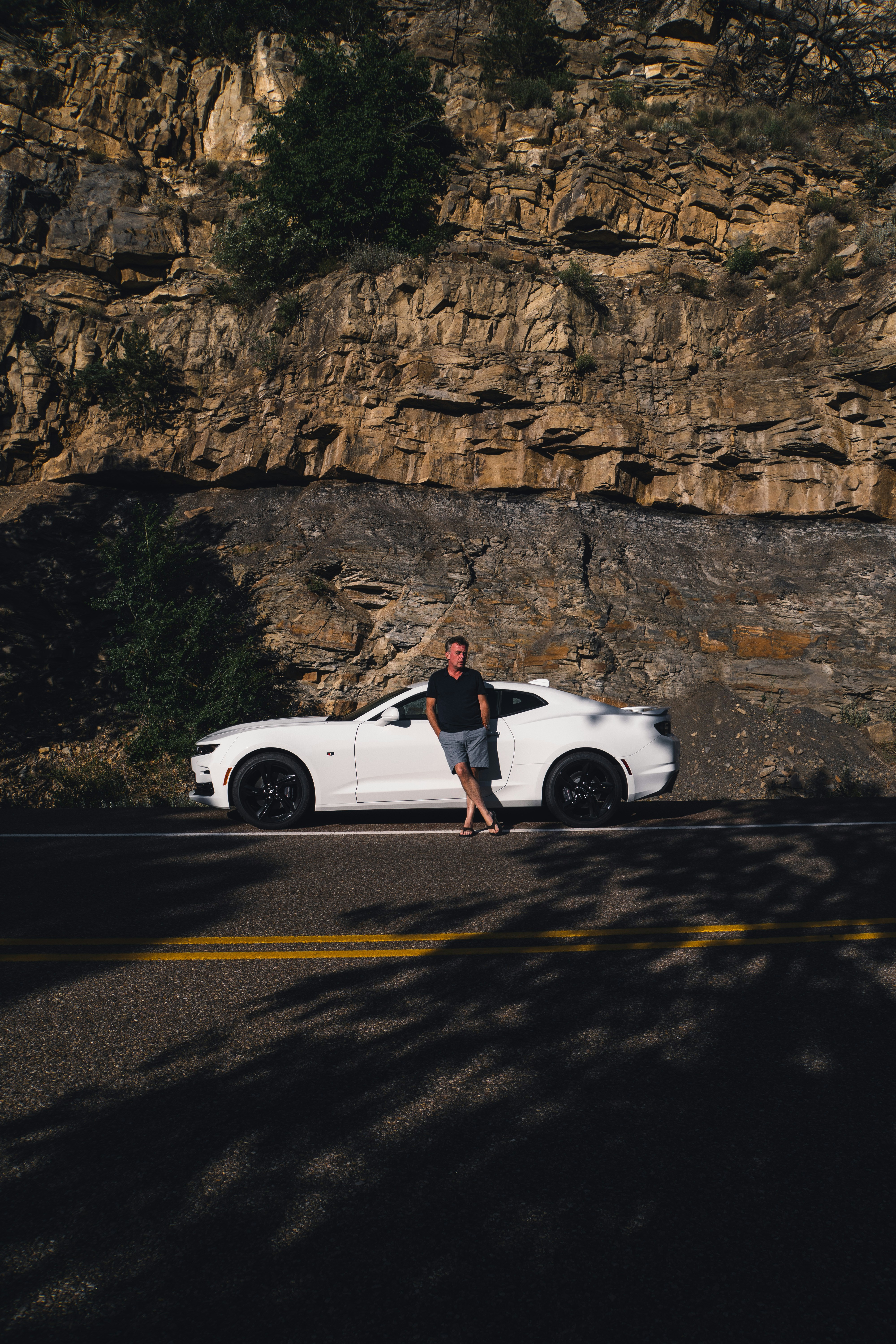 Sunlit Cliffside Pause: A White Sports Car and a Leaning Figure on a Mountain Road