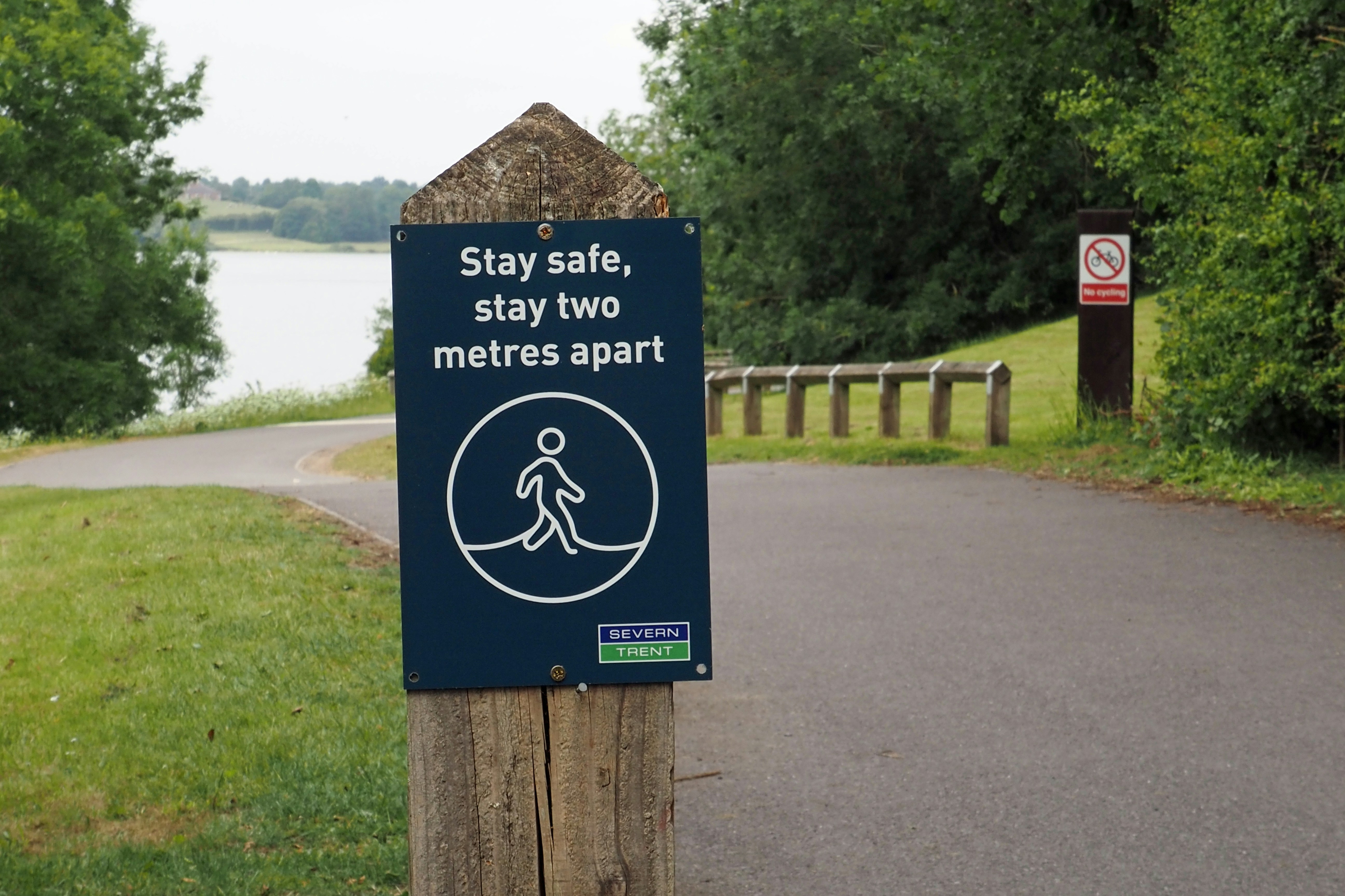Blue and white sign advising social distancing on a lakeside path with lush greenery.