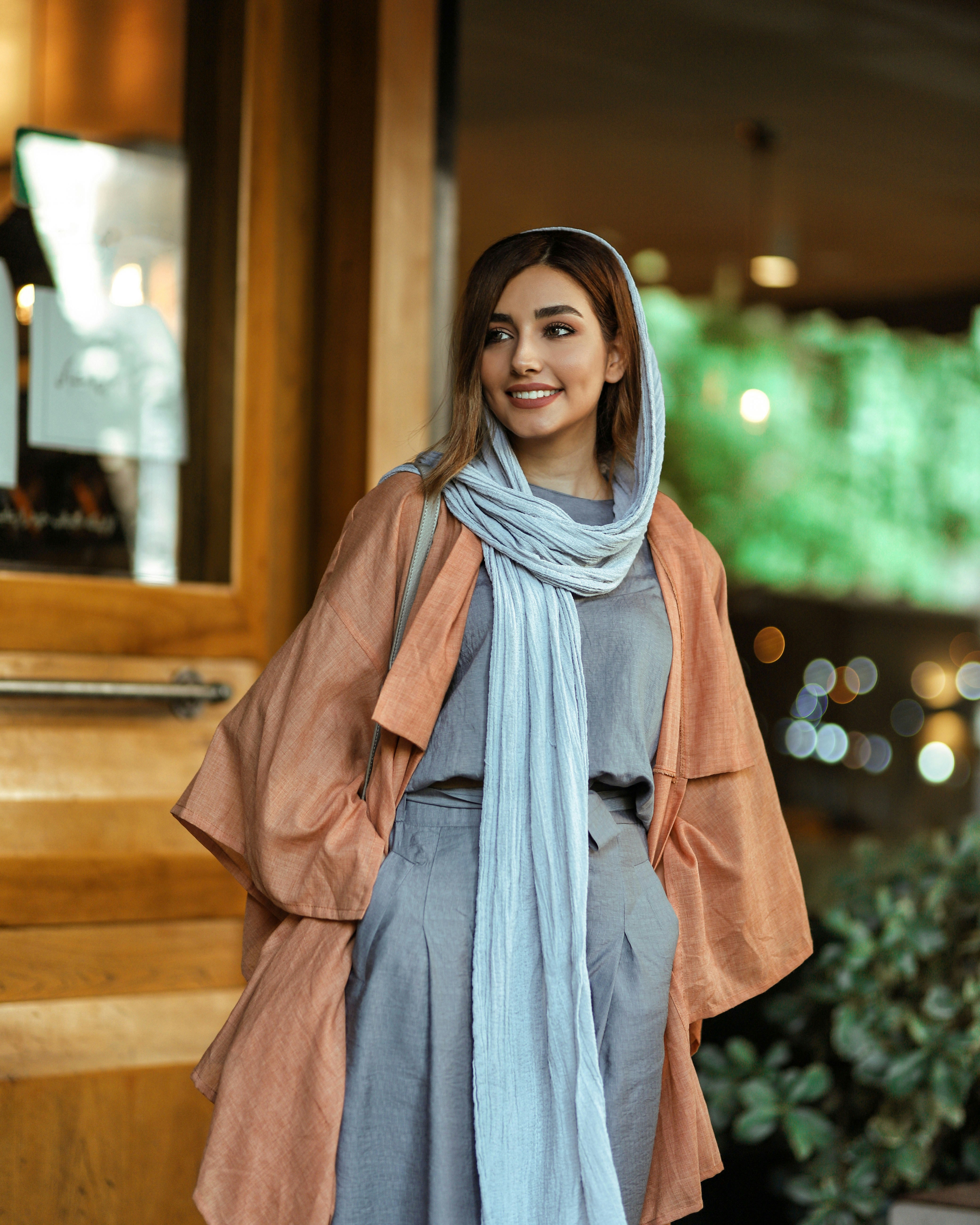 A woman dressed in a flowing orange coat and light blue scarf stands near a wooden entrance, exuding a sense of urban sophistication. Soft bokeh lights create a warm atmosphere.