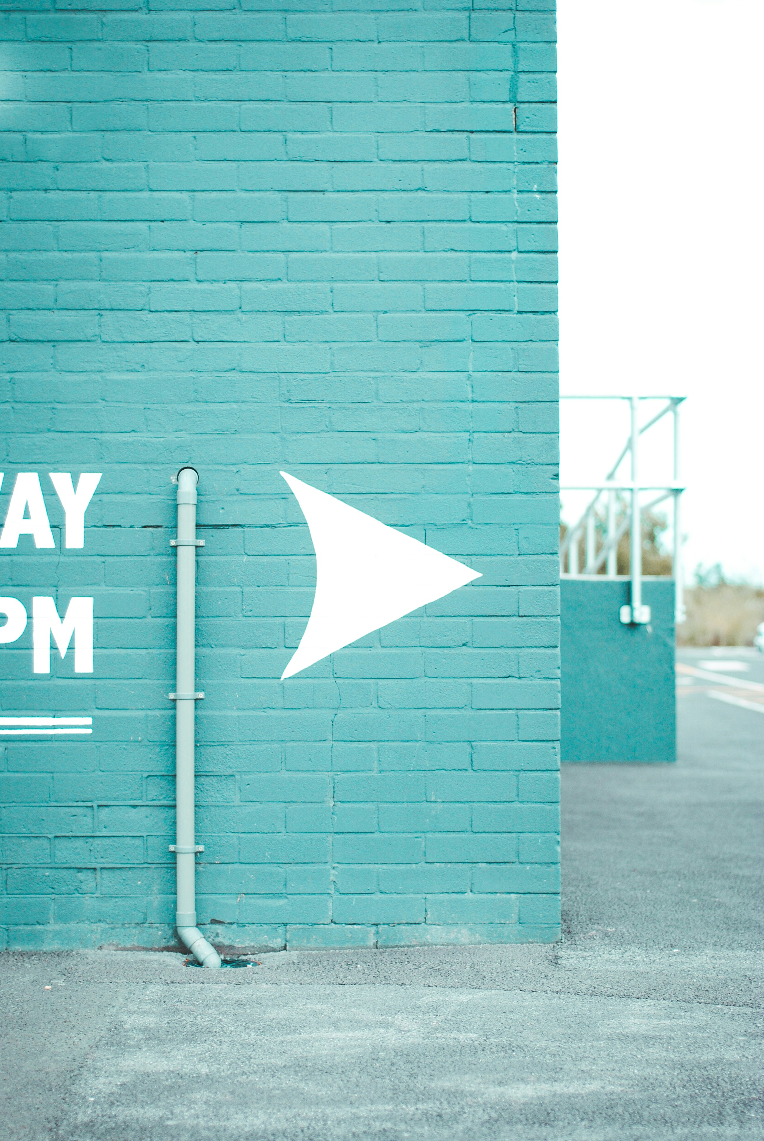 A teal brick wall features a bold white arrow and text indicating direction, complemented by a gray sidewalk. The composition emphasizes urban signage.