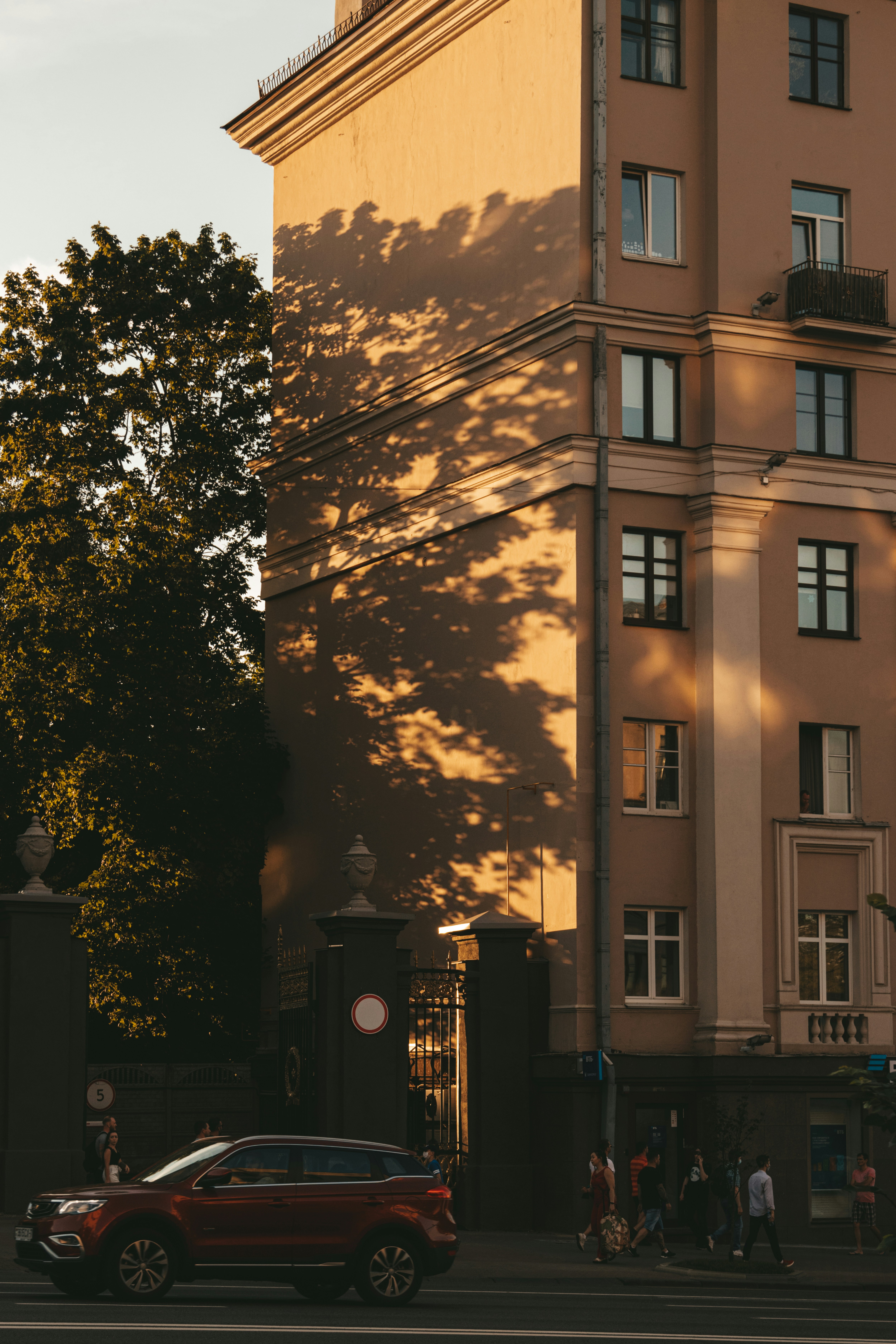 The interplay of light and shadow on an urban building highlights the architectural details against a backdrop of lush greenery. A red car moves past, adding a dynamic element to the scene.