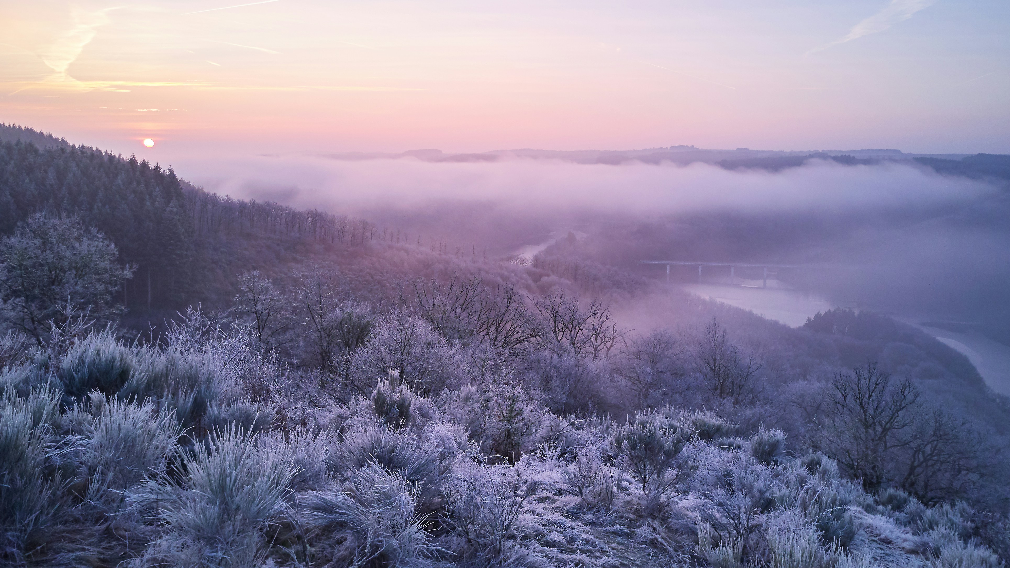 Sunrise over a frosty landscape with mist enveloping a distant valley and bridge.
