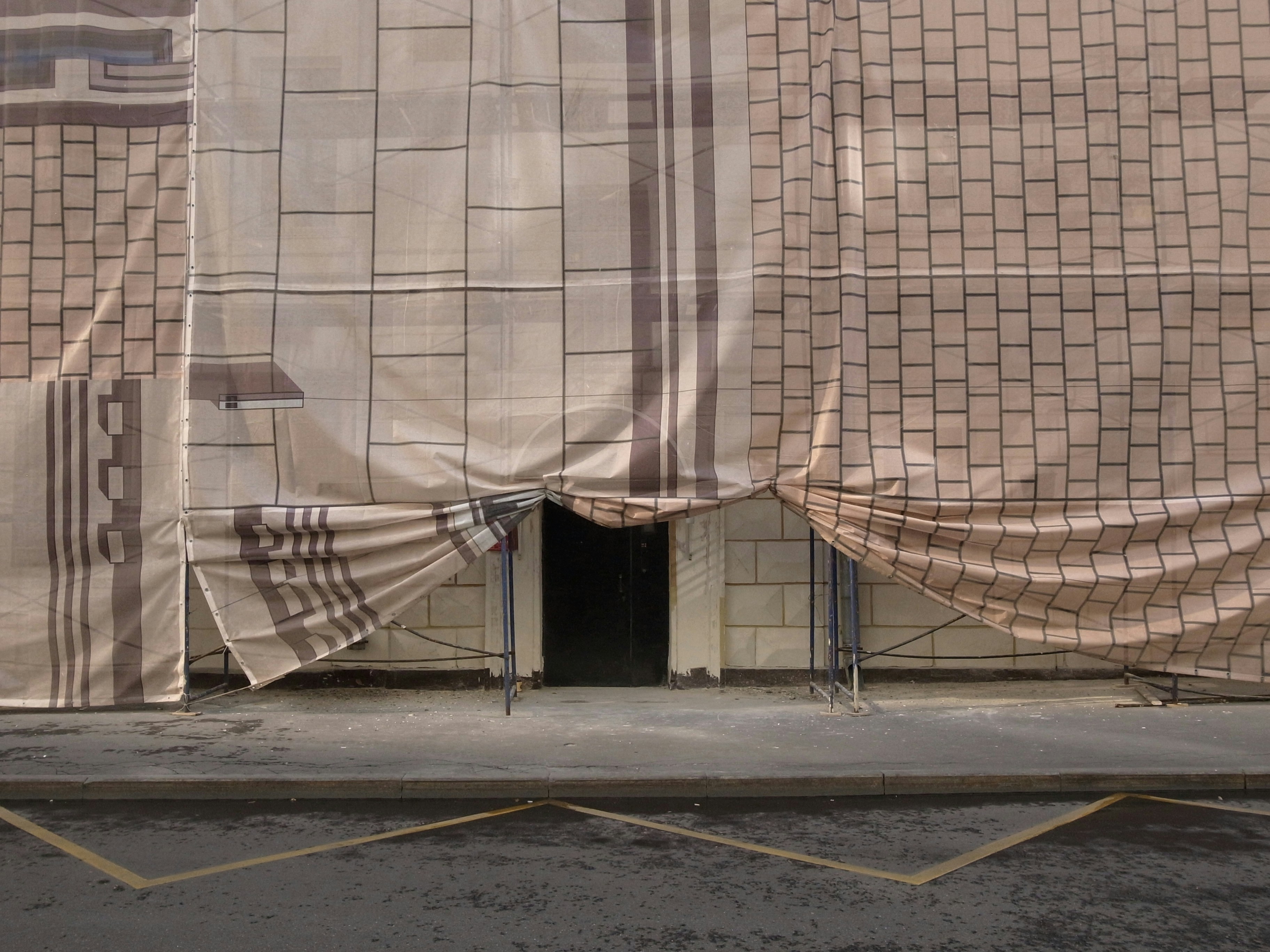 Construction site obscured by patterned tarpaulin, hinting at architectural changes behind the fabric. The entrance remains partially visible.
