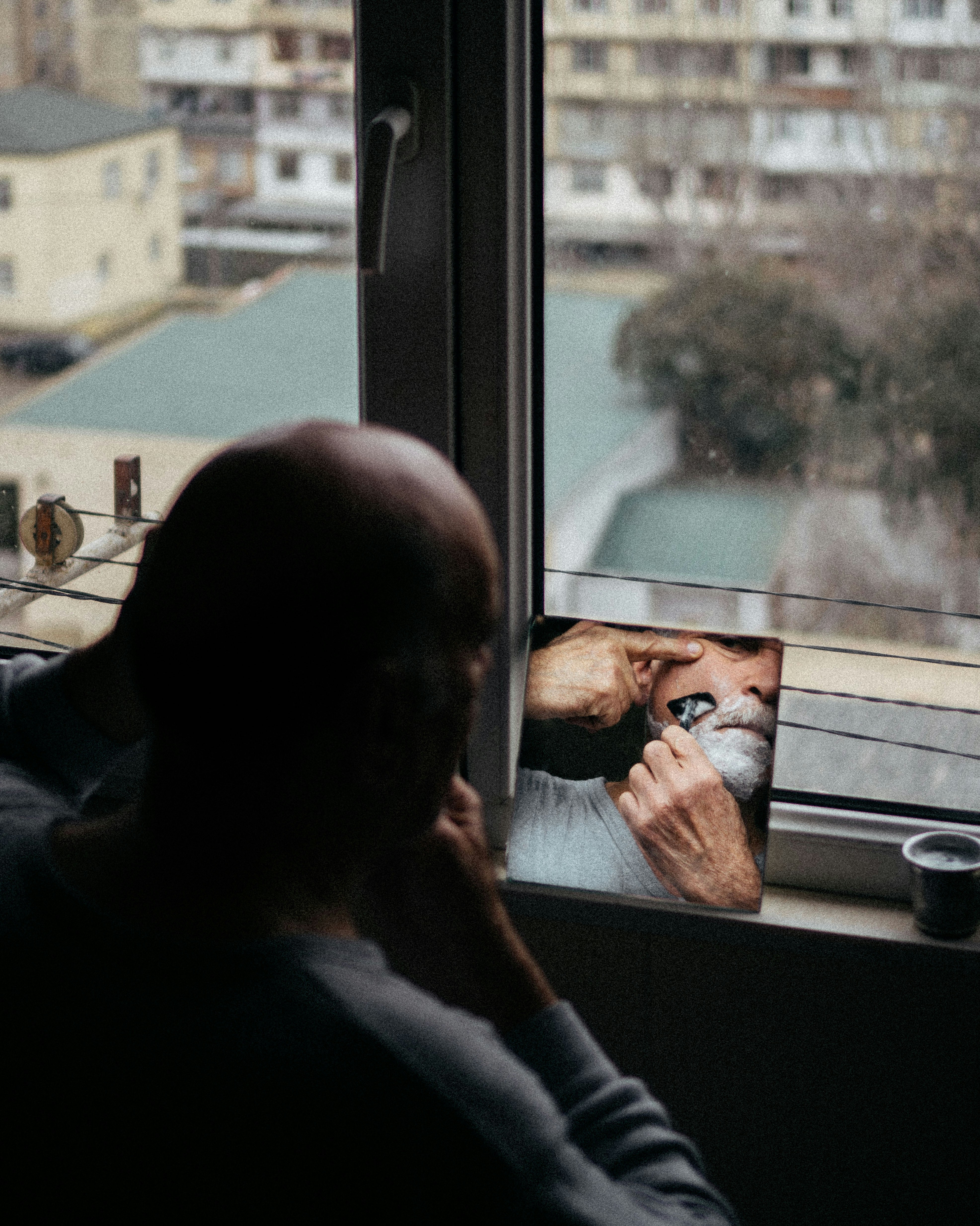 An elderly man grooming himself, reflected in a mirror on a windowsill, with a view of the outside world in the background.