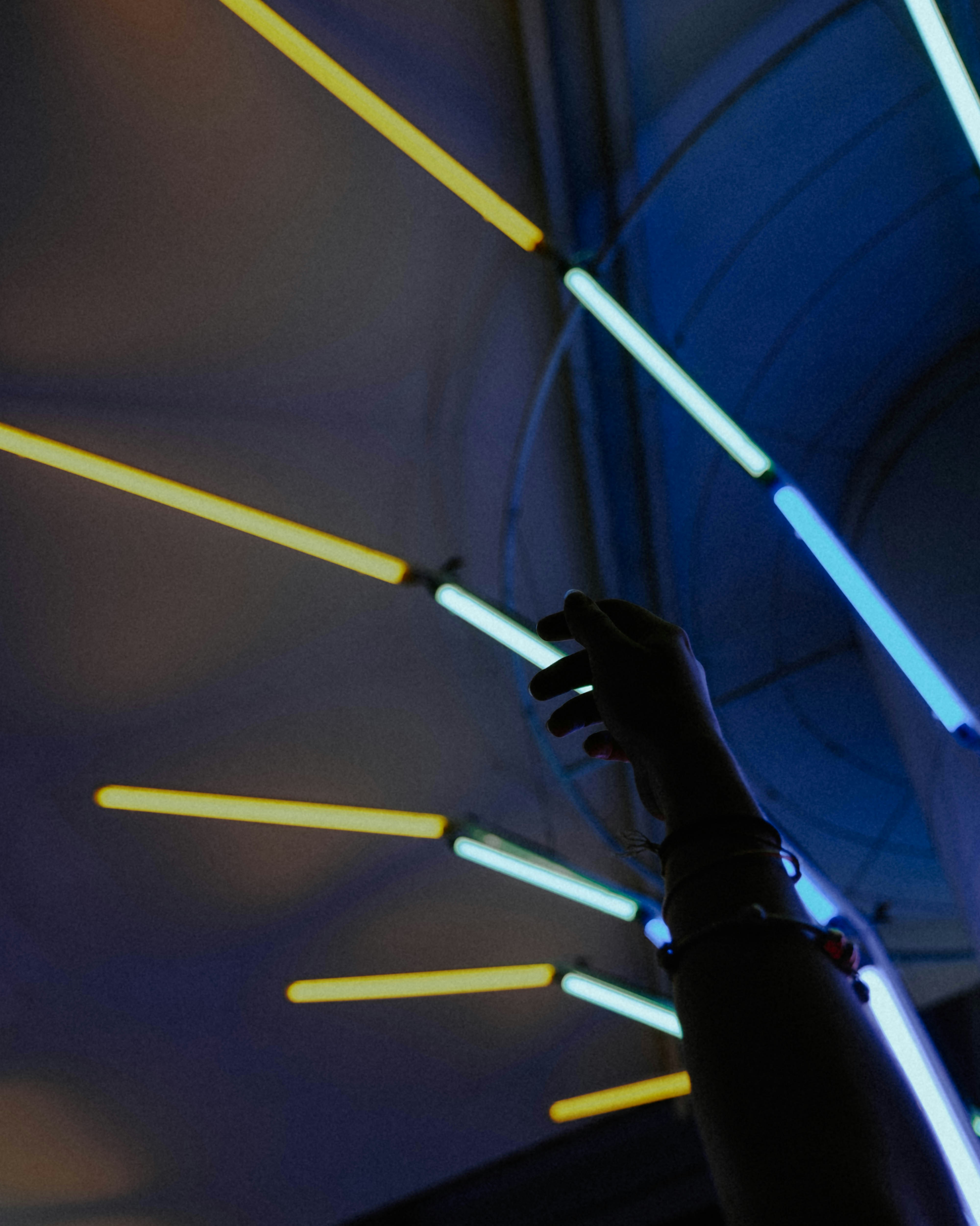 A hand reaches toward glowing neon lights in a darkened space, creating a dynamic interplay of color and shadow.