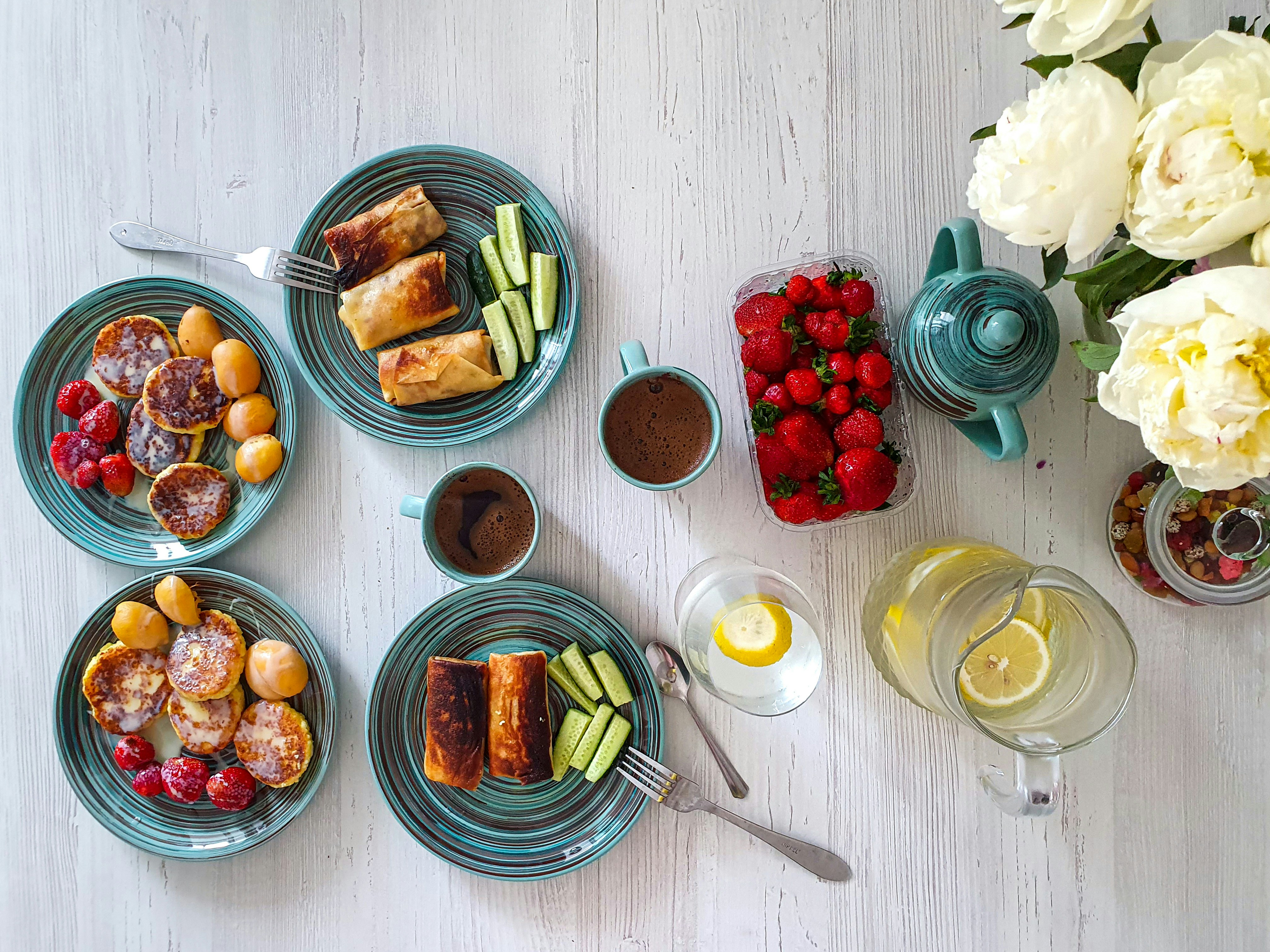 Overhead view of a bright breakfast spread featuring pastries, berries, and citrus drinks arranged on turquoise plates, with white flowers nearby.