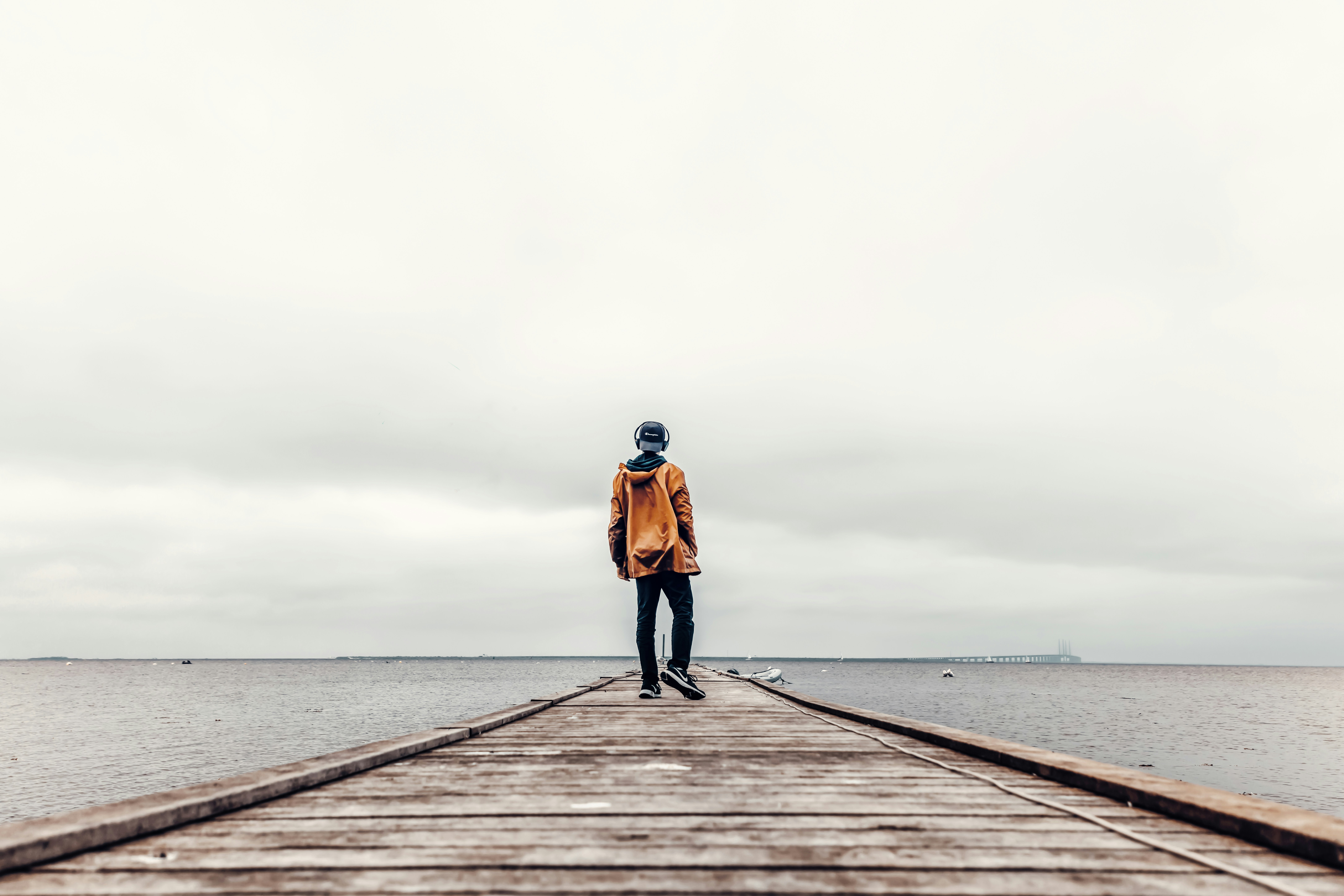 A lone figure stands on a weathered pier, gazing into the overcast horizon where water meets sky.