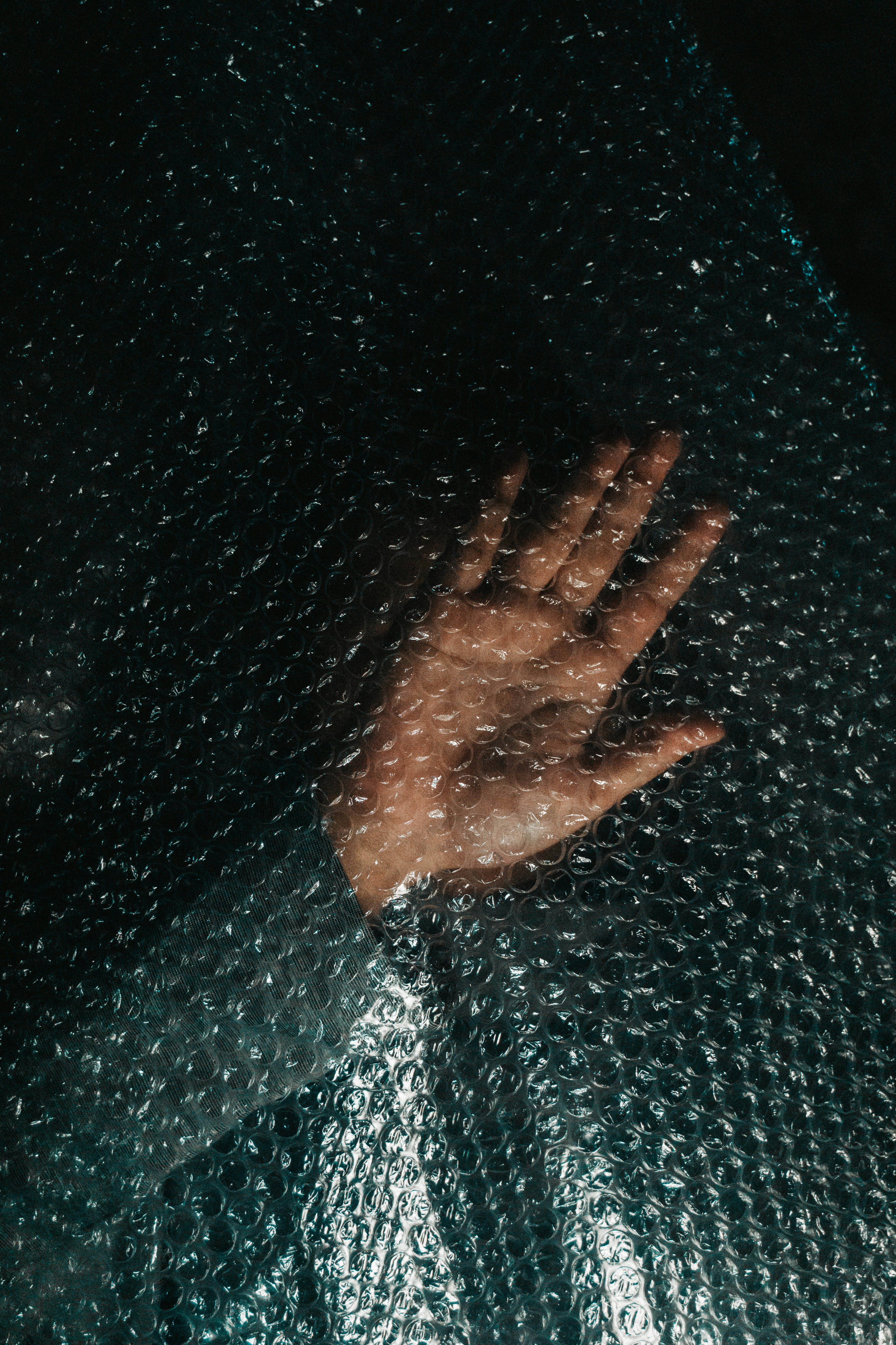 Close-up photograph of a hand pressed against bubble wrap, catching light through the bubbles. The texture and depth of the translucent plastic are highlighted.