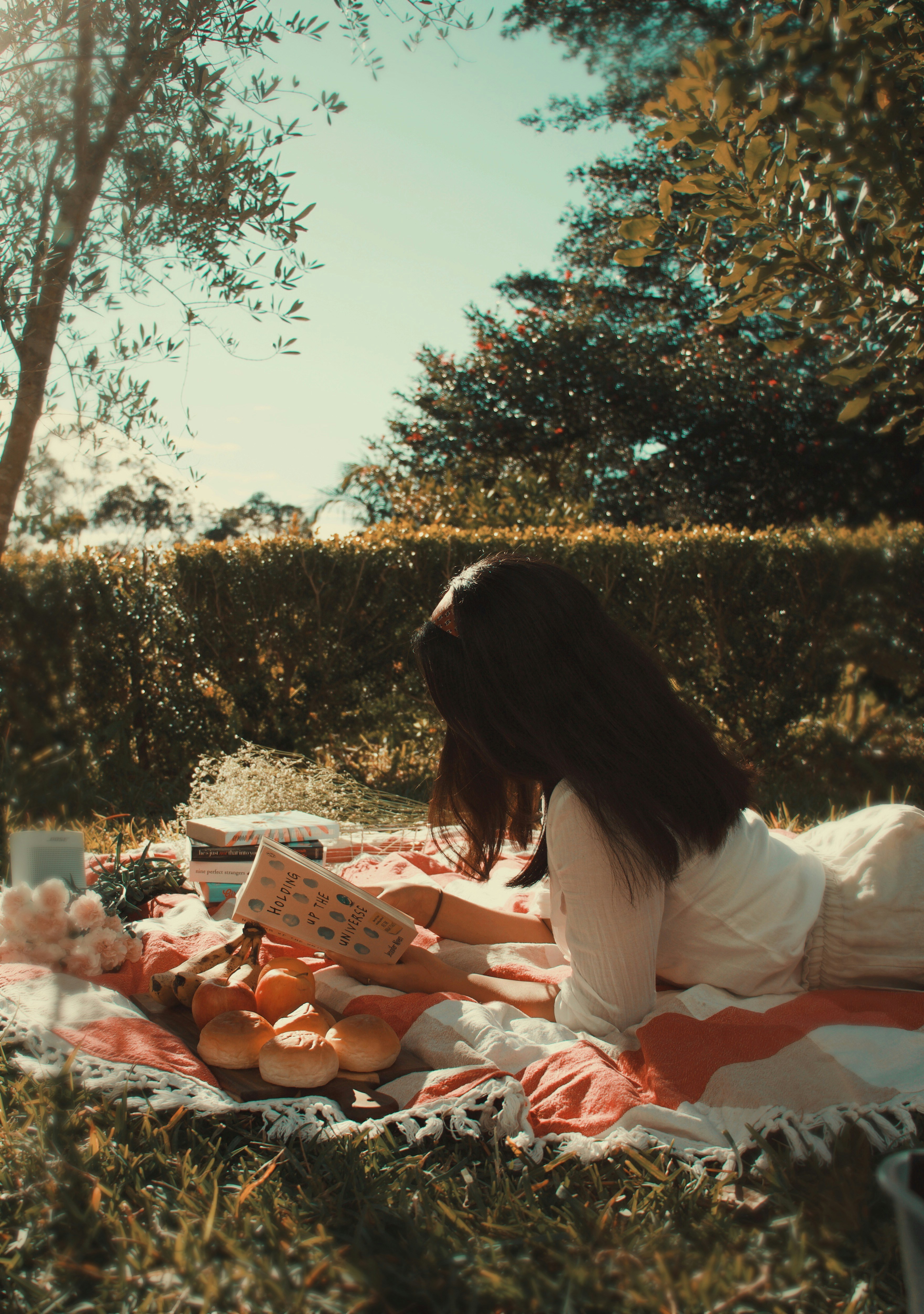 Woman reading a book while lounging on a picnic blanket surrounded by fruits and flowers in a lush garden setting.