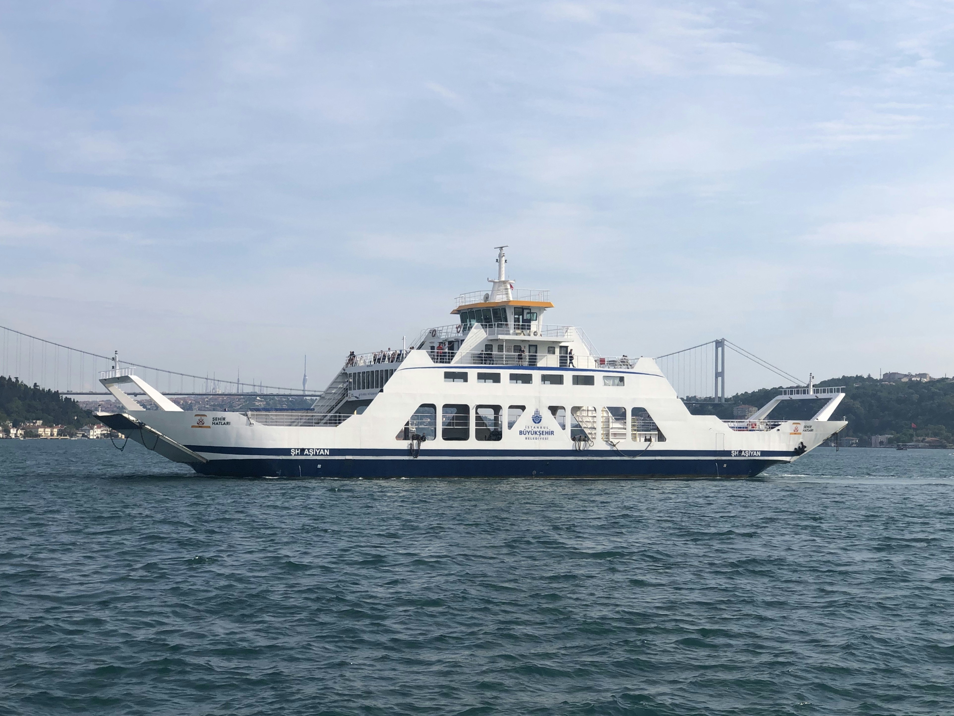 A large ferry navigating the waters of the Bosphorus, with a bridge in the background and passengers enjoying the journey.