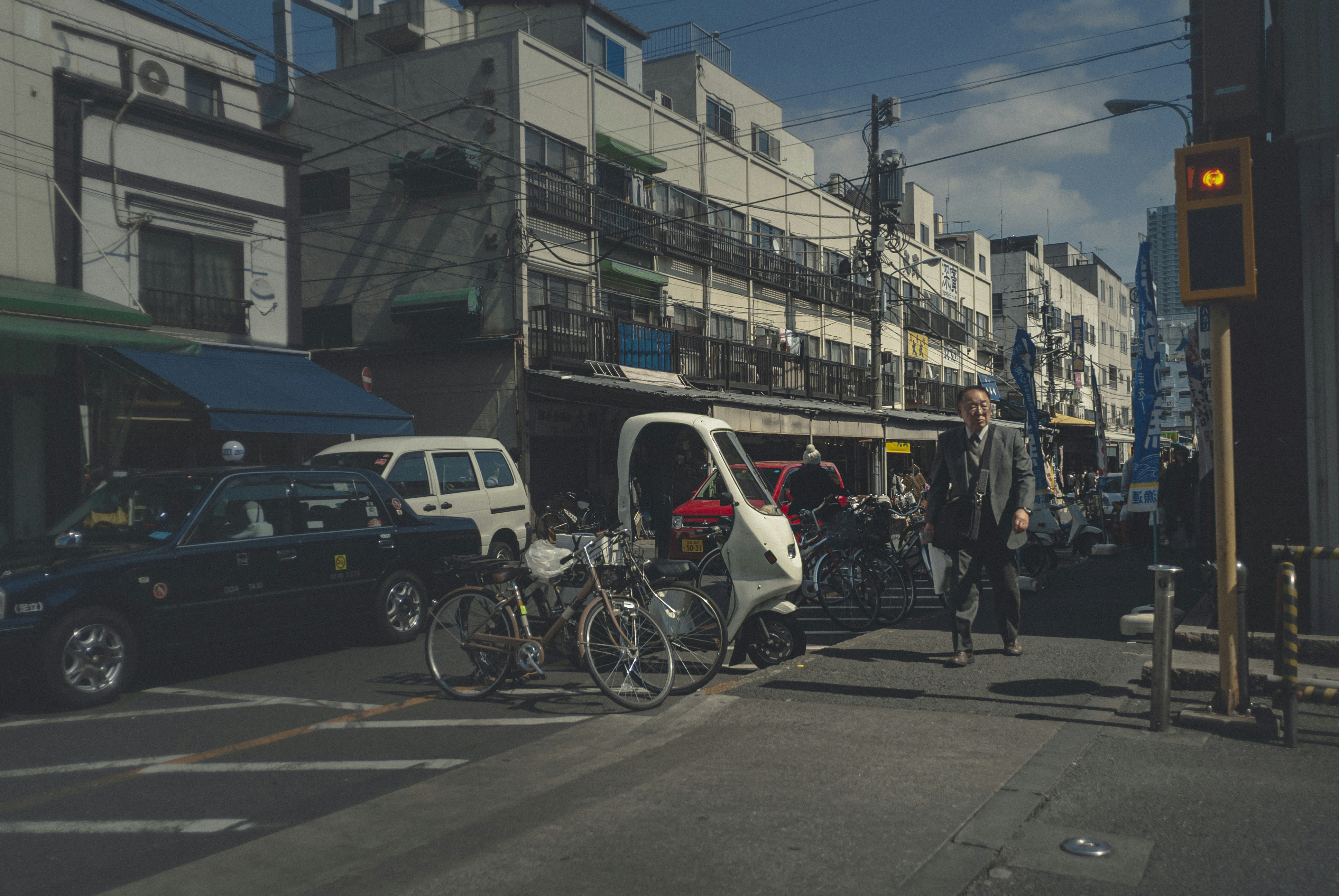 Busy street scene featuring a man in a suit walking past parked cars and bicycles, with buildings lining the background. The urban environment captures the essence of daily life.
