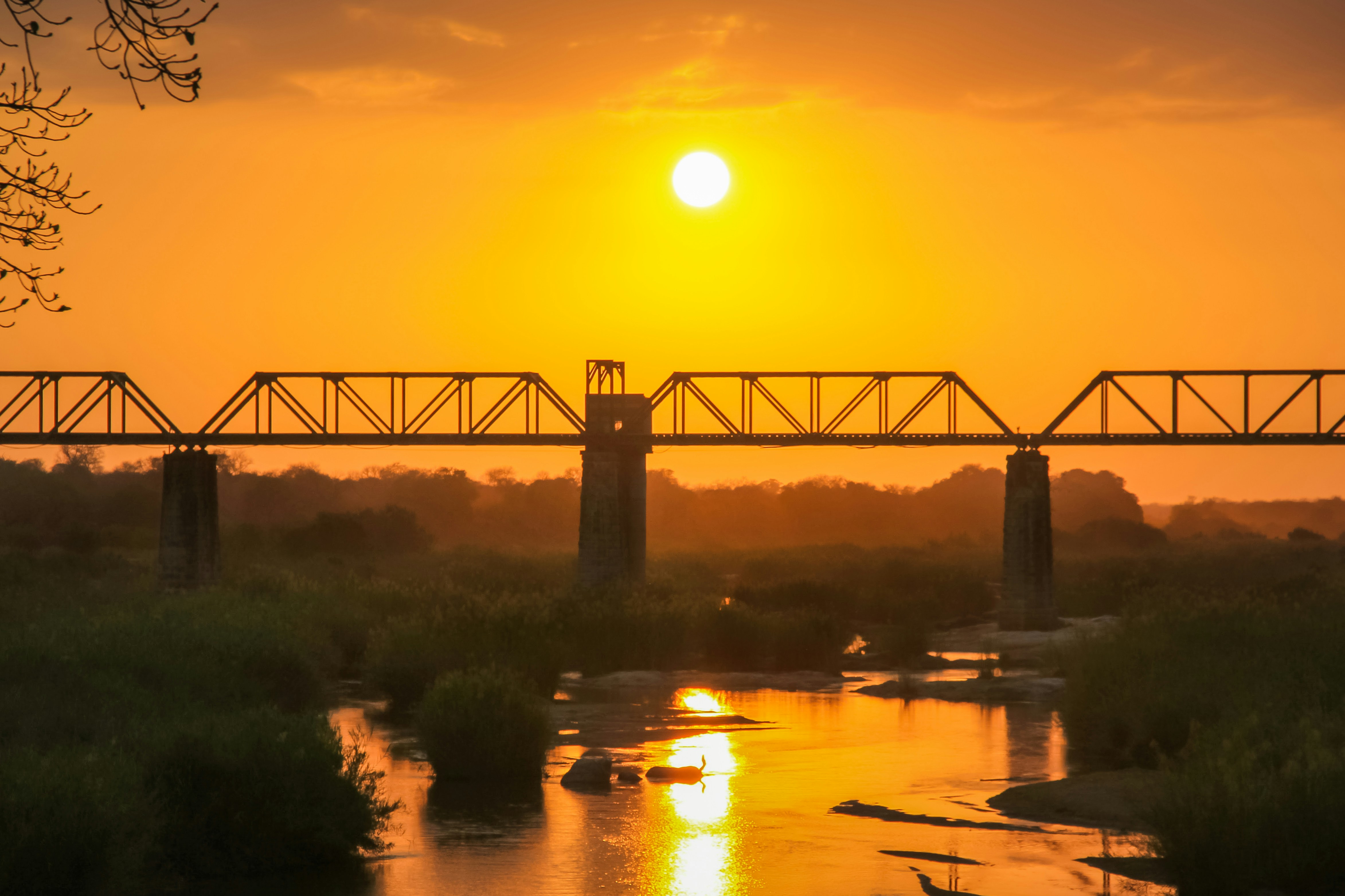 Silhouetted bridge spans across a tranquil river as the sun sets, casting golden reflections on the water's surface.