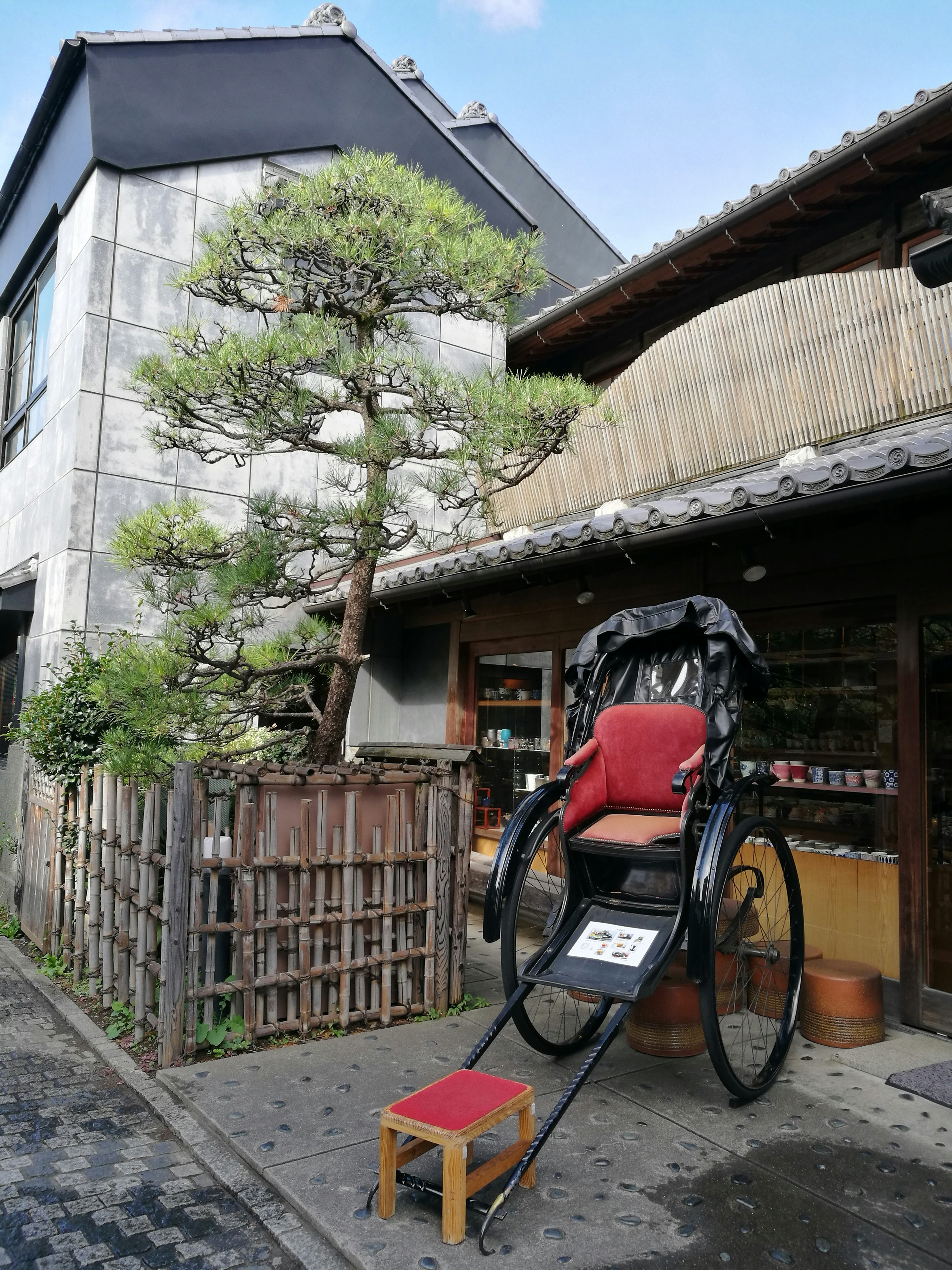 Historic rickshaw beside a traditional building and bonsai tree, under a clear blue sky.