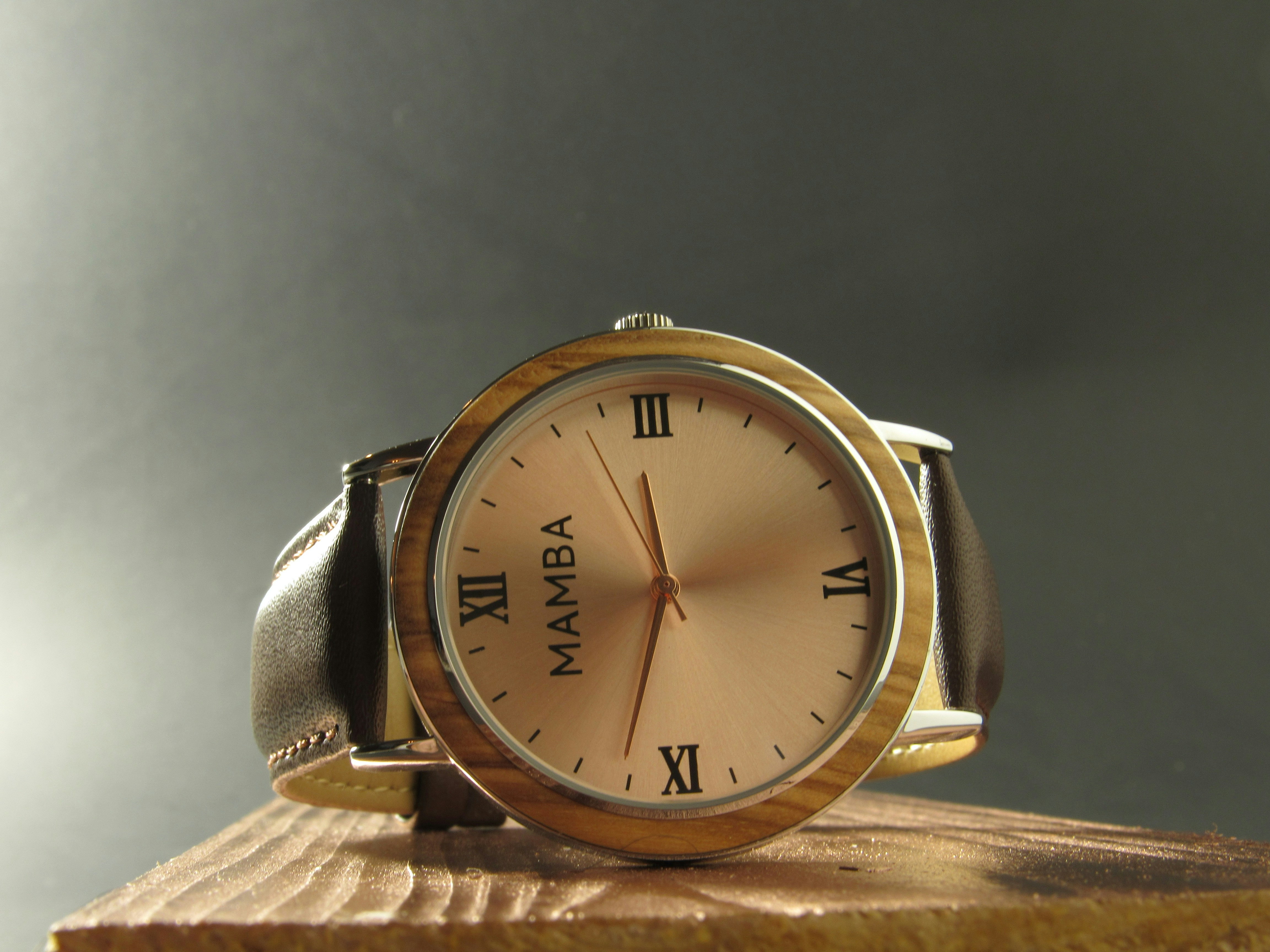 A close-up of a Mamba watch featuring a minimalist design with Roman numerals and a brown leather strap resting on a wooden surface.