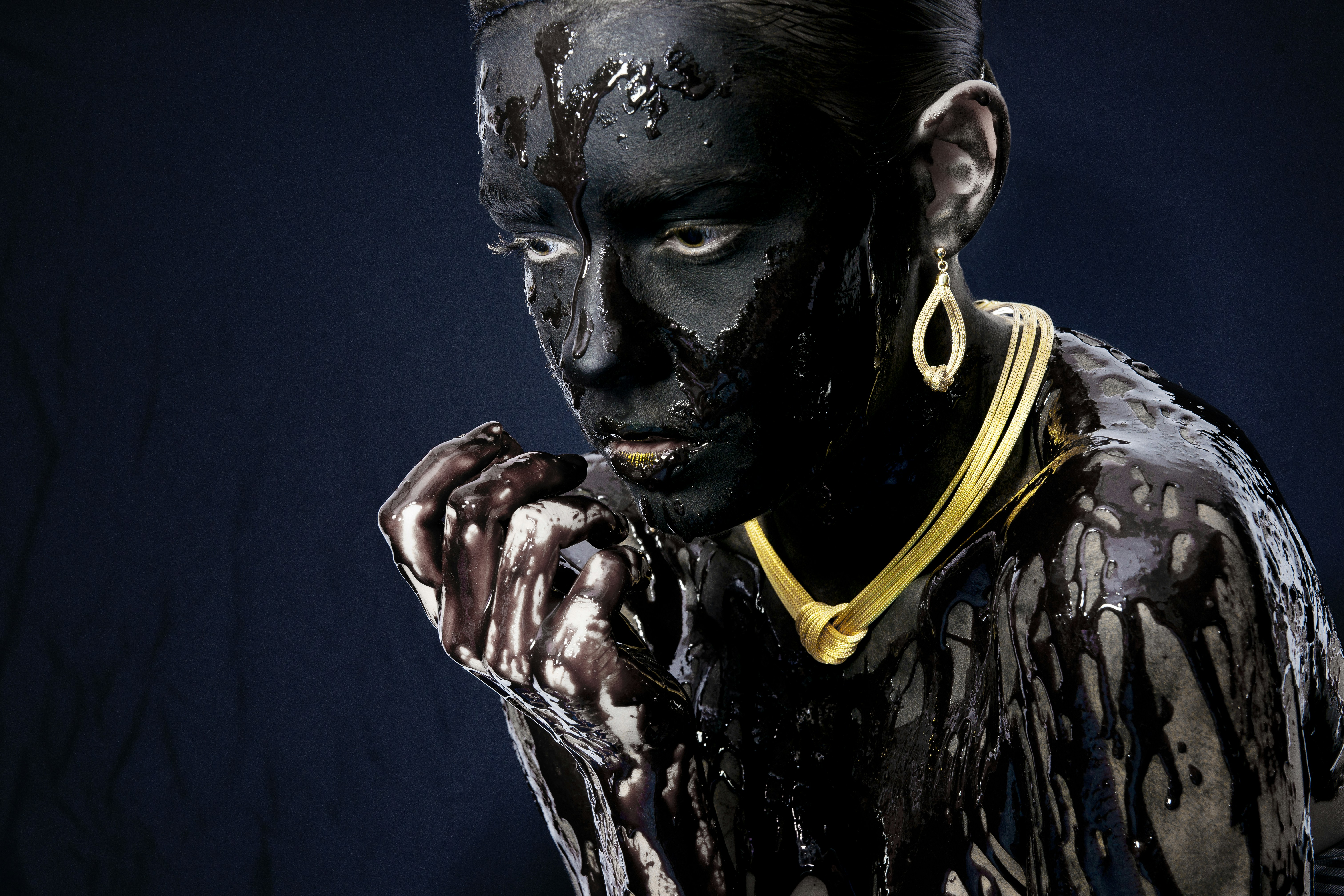 Studio portrait photograph of a person with glossy black body paint and gold jewelry against a dark backdrop.
