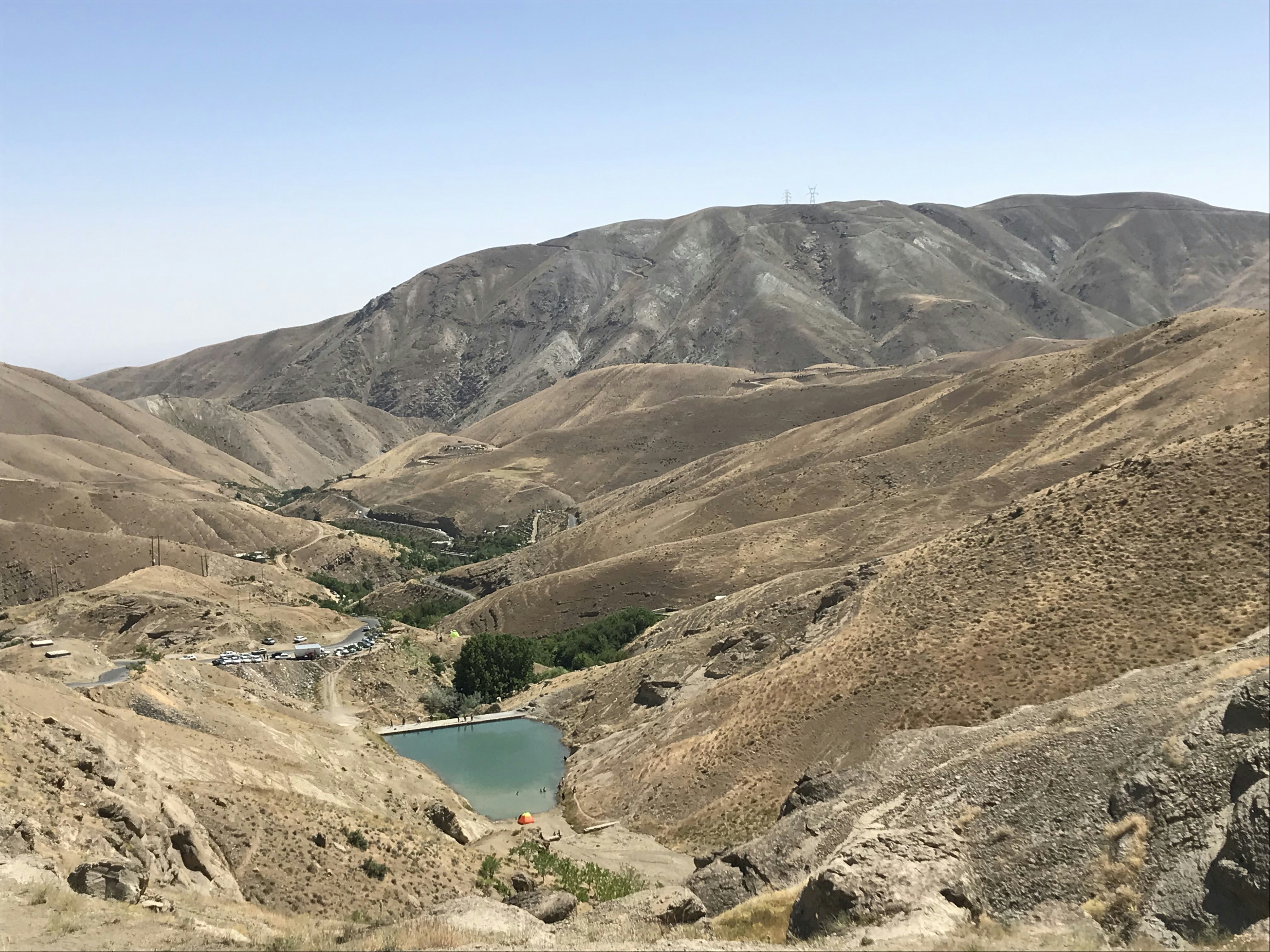 A serene water body nestled within dry hills, surrounded by rugged terrain and sparse vegetation. The scene captures the contrast between the lush oasis and the barren landscape.