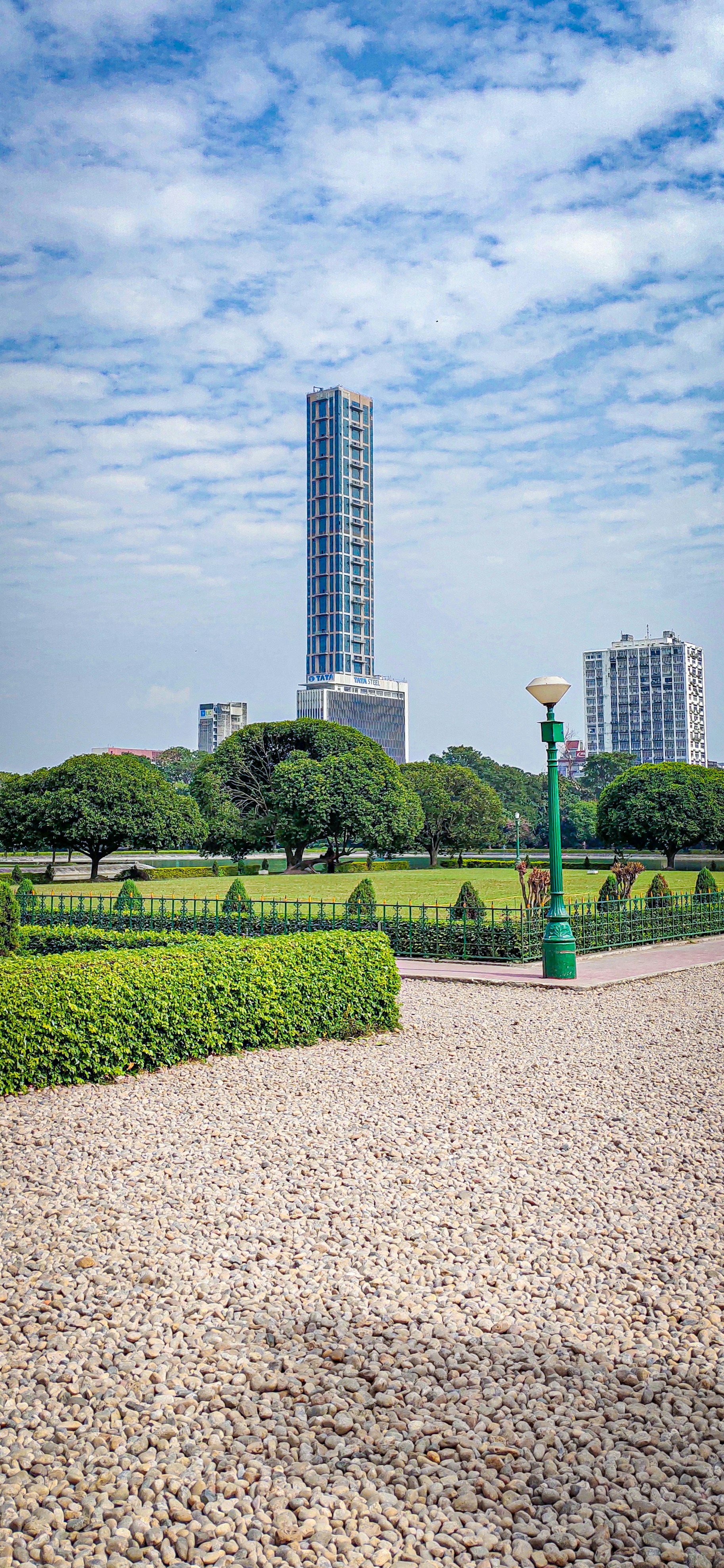 A photograph captures a slender high-rise behind a manicured park with a gravel foreground and a bright sky with scattered clouds.