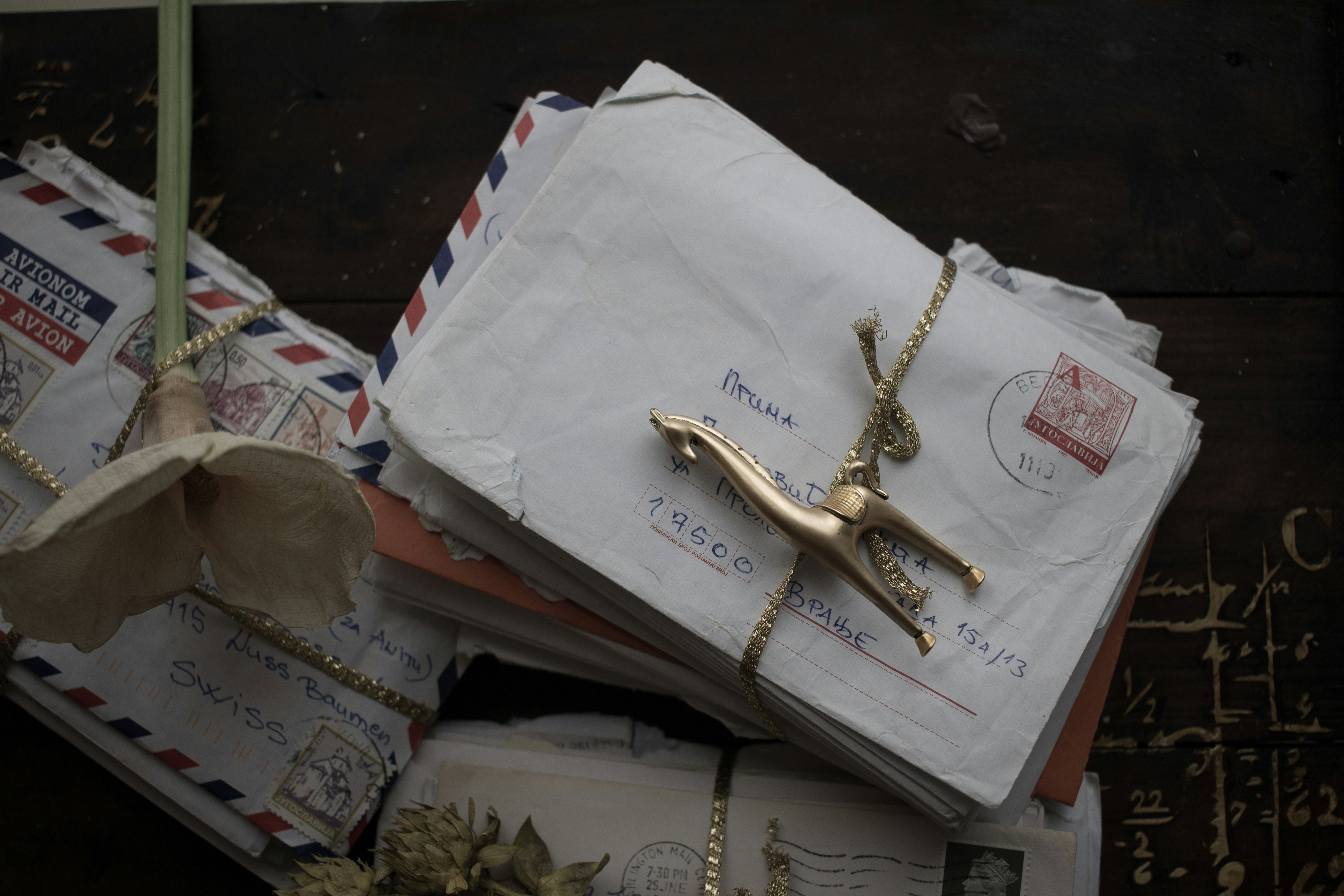 A stack of vintage airmail envelopes tied with ribbon sits atop a wooden surface, with a delicate metal bird charm resting on the top envelope. The scene conveys a sense of romance and correspondence.