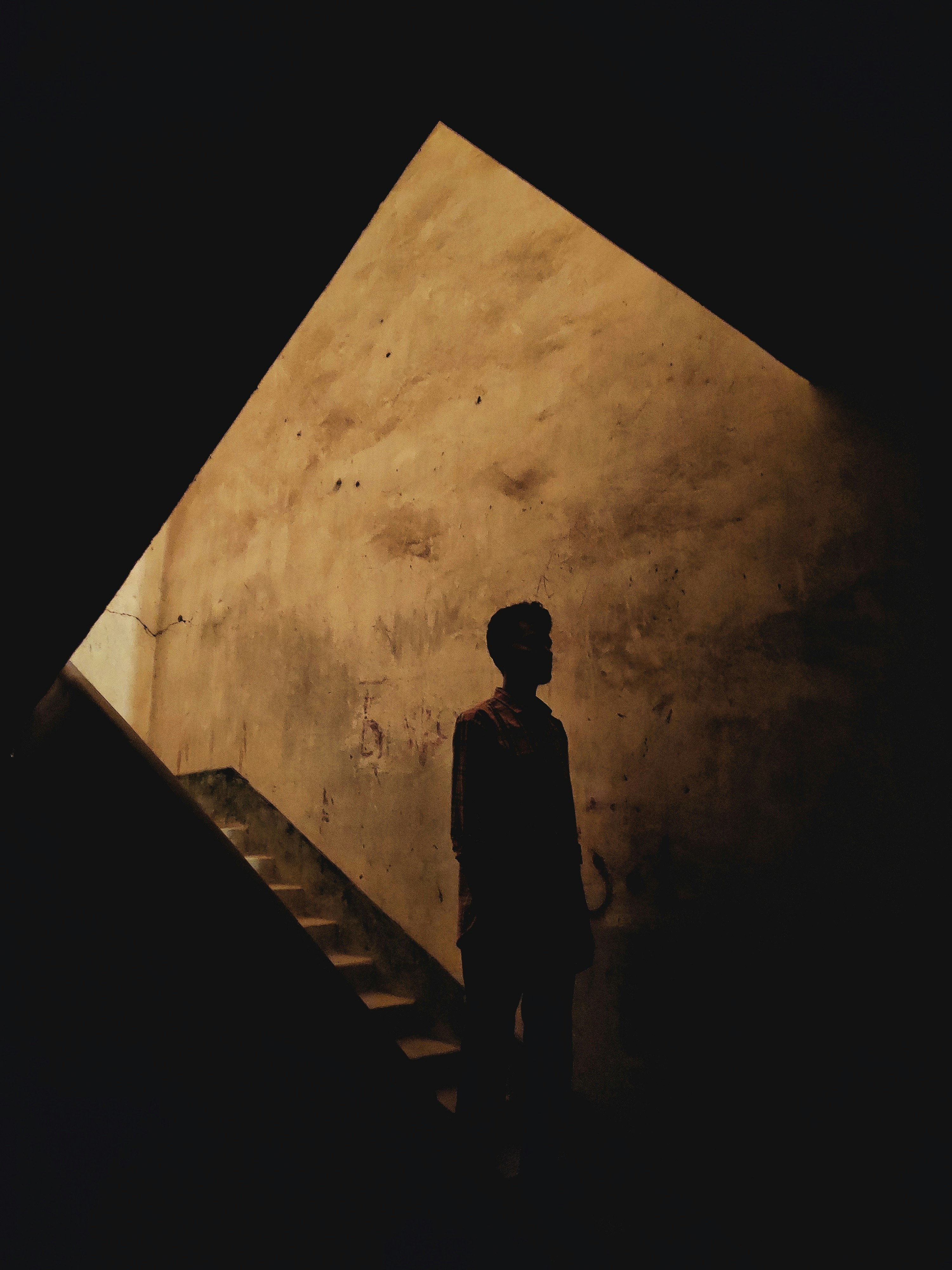 Silhouette of a figure standing on stairs against a textured wall illuminated by soft light.