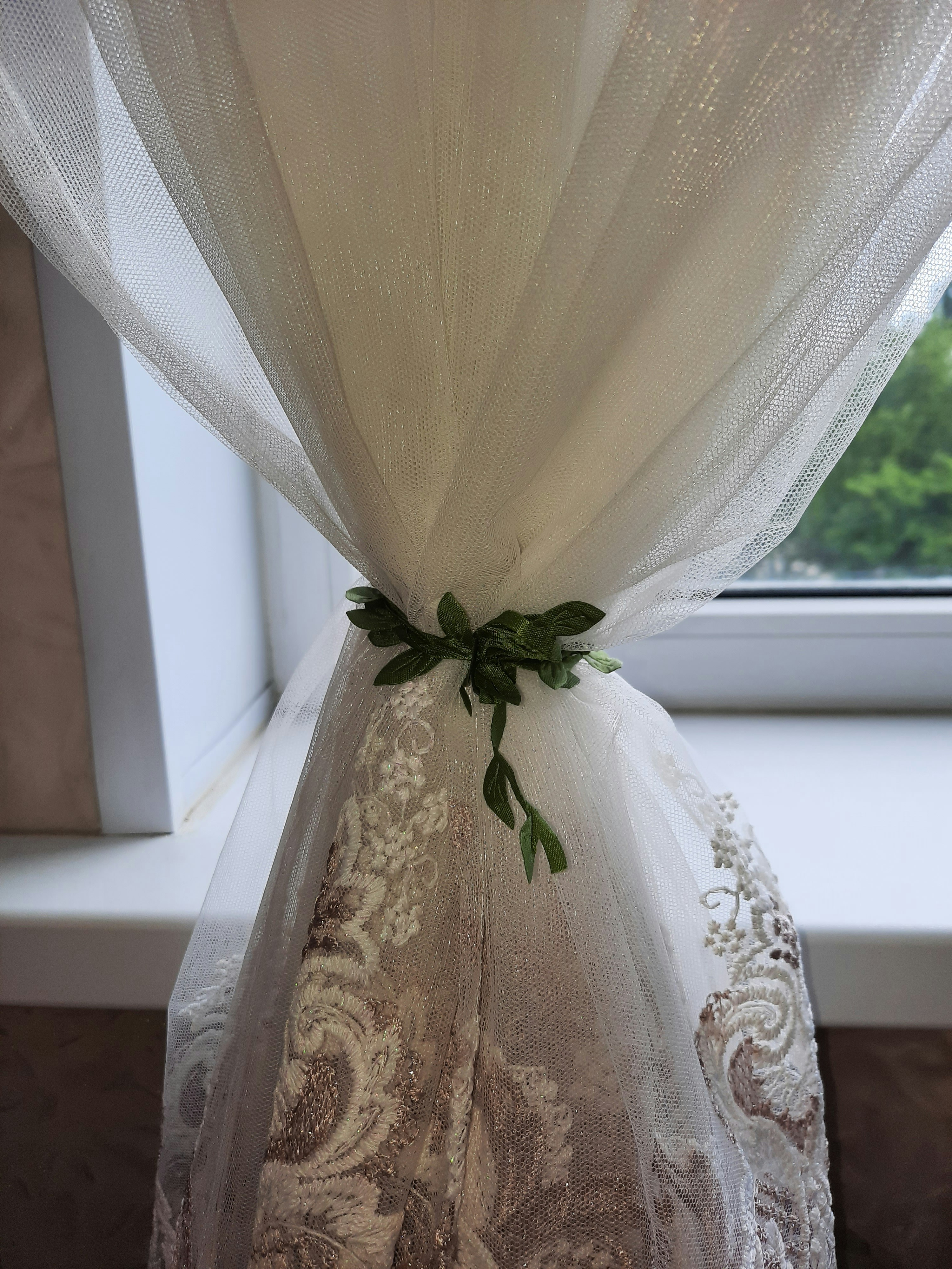 Delicate sheer curtain tied with greenery, framing a window view. The intricate lace details add a touch of sophistication.