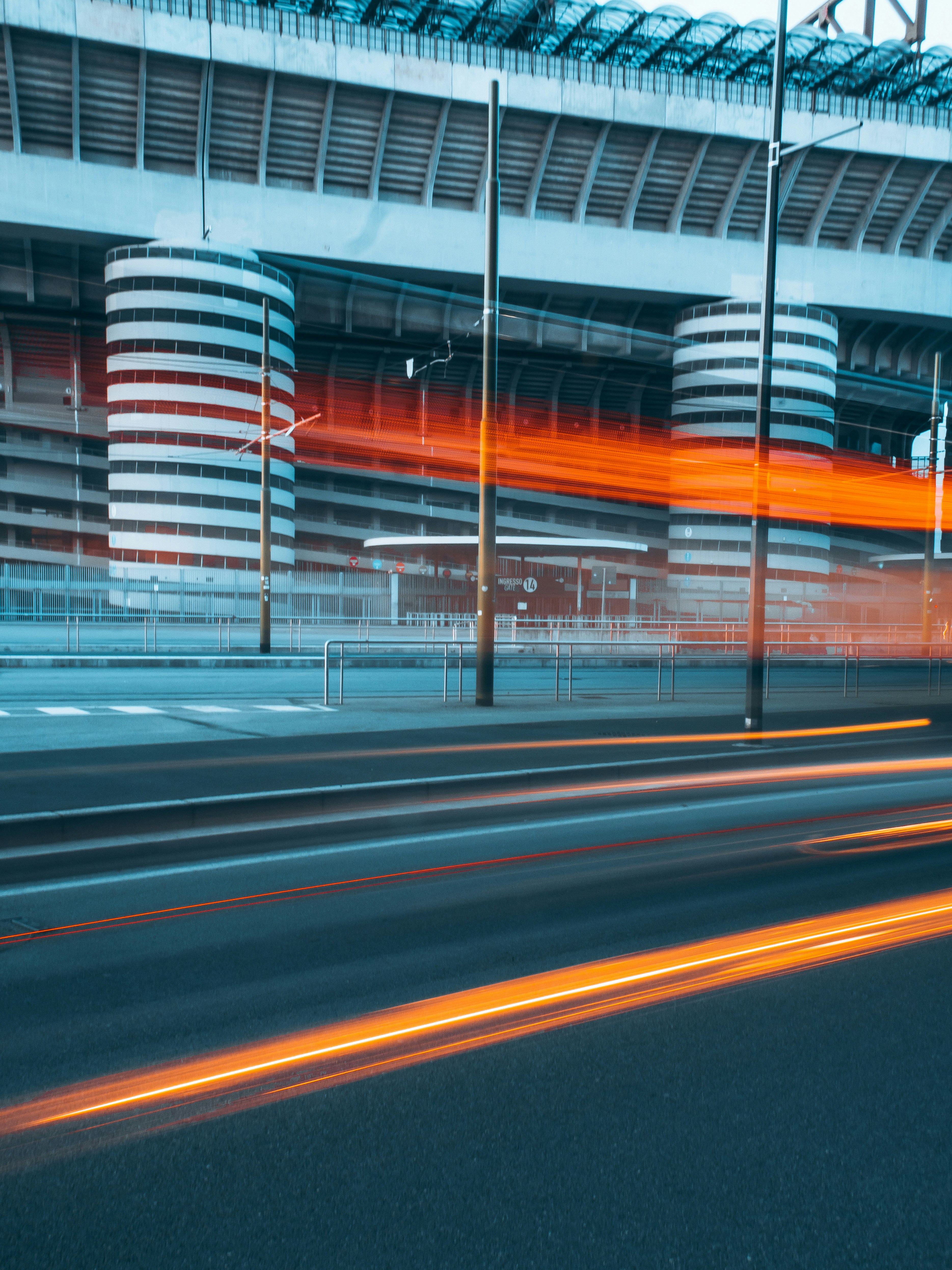 Long-exposure photograph of a city street beneath an elevated viaduct, with neon red and orange light trails slicing across the road and striped cylindrical columns in the background.