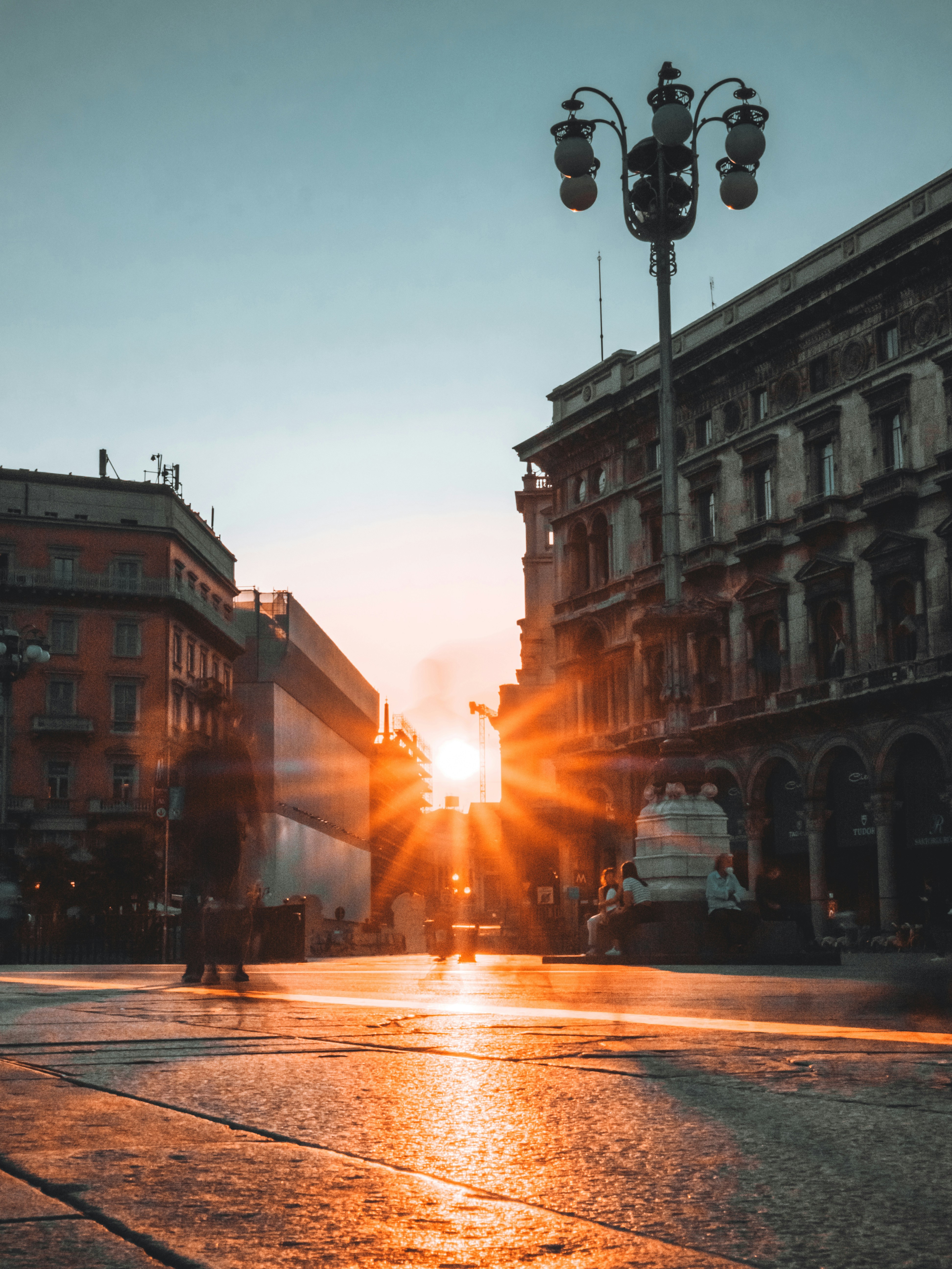 A photograph captures a sunburst between historic buildings lighting a cobblestone plaza at dusk, with motion-blurred pedestrians and glowing reflections.