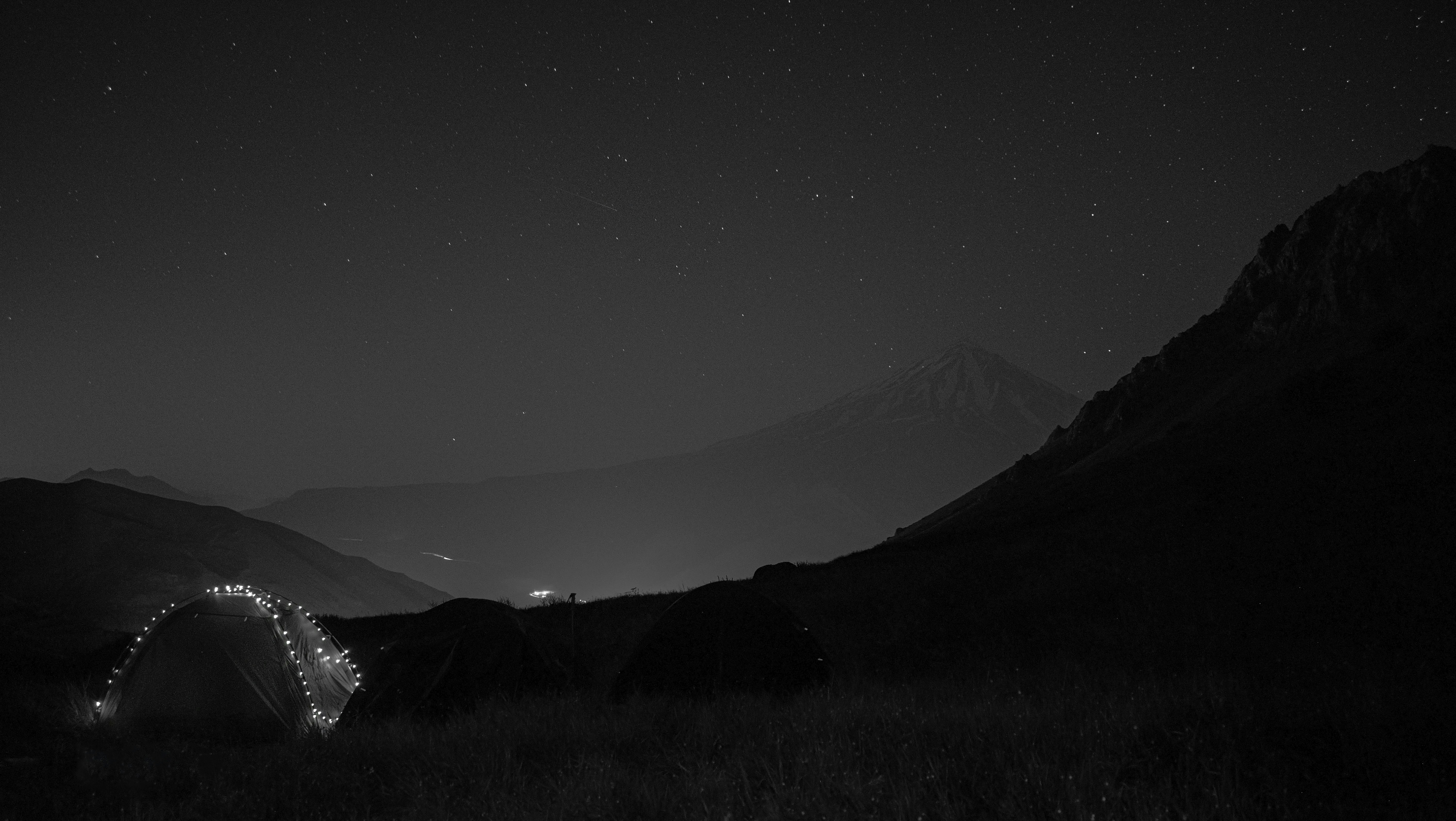 Grayscale photo of a tent adorned with fairy lights beneath a starry sky in a mountainous landscape.
