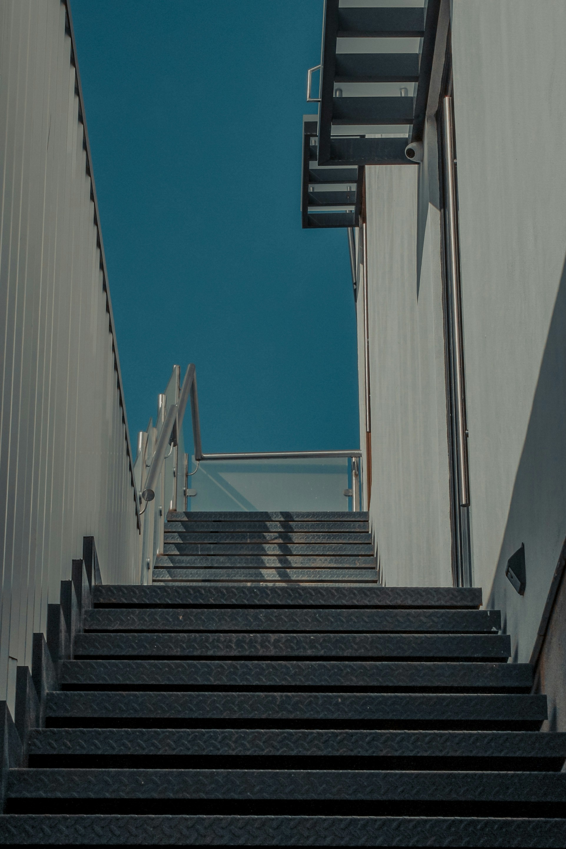 Metal staircase leading upwards towards a clear blue sky, framed by sleek white walls and a balcony.