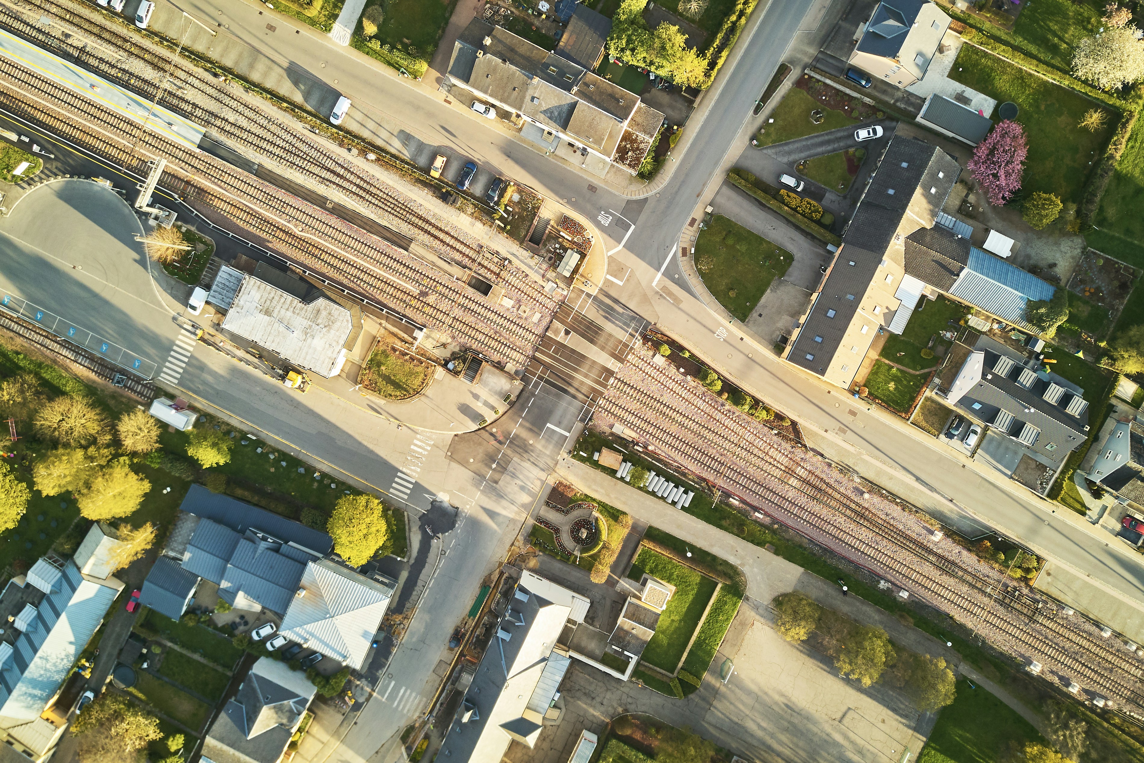 Aerial view of a railway crossing intersecting with a residential street, showcasing the layout of buildings and greenery in the surrounding area.