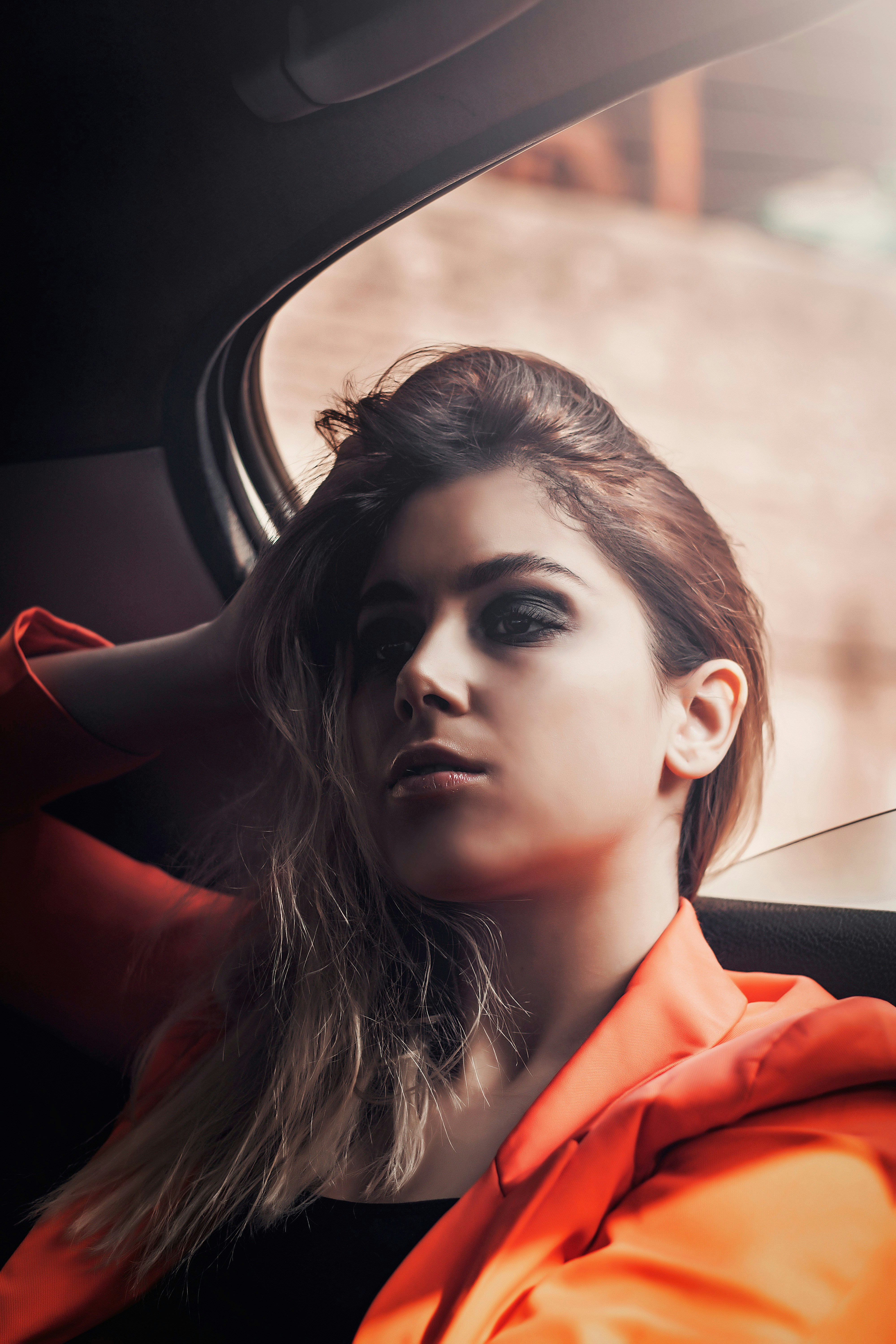 A young woman with striking features poses thoughtfully in a car, her vibrant orange blazer contrasting with the muted background. The soft lighting enhances her contemplative expression.