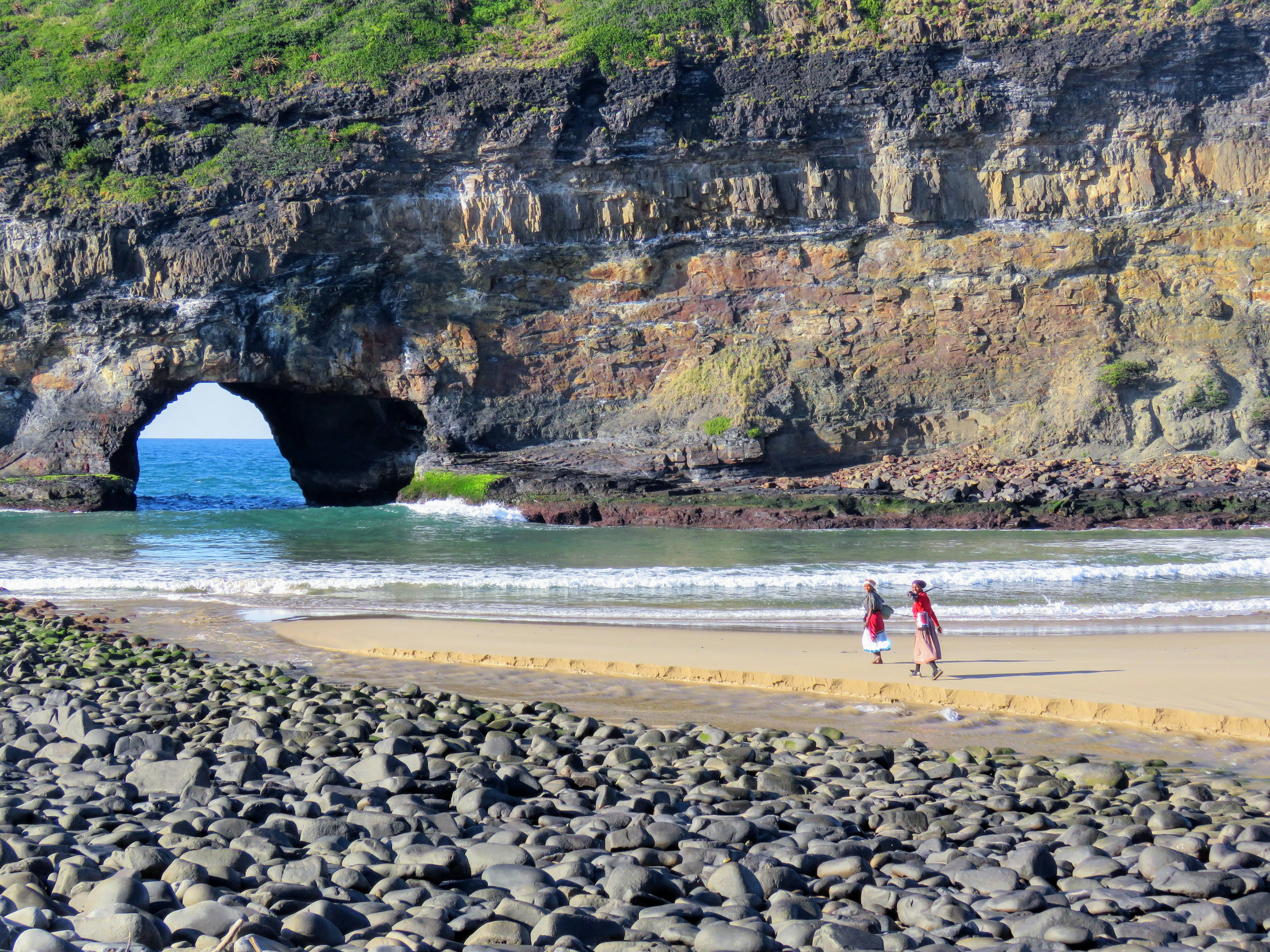 Two figures walking along a sandy beach, framed by a dramatic rock arch and gentle ocean waves. The scene captures the serene beauty of coastal landscapes.