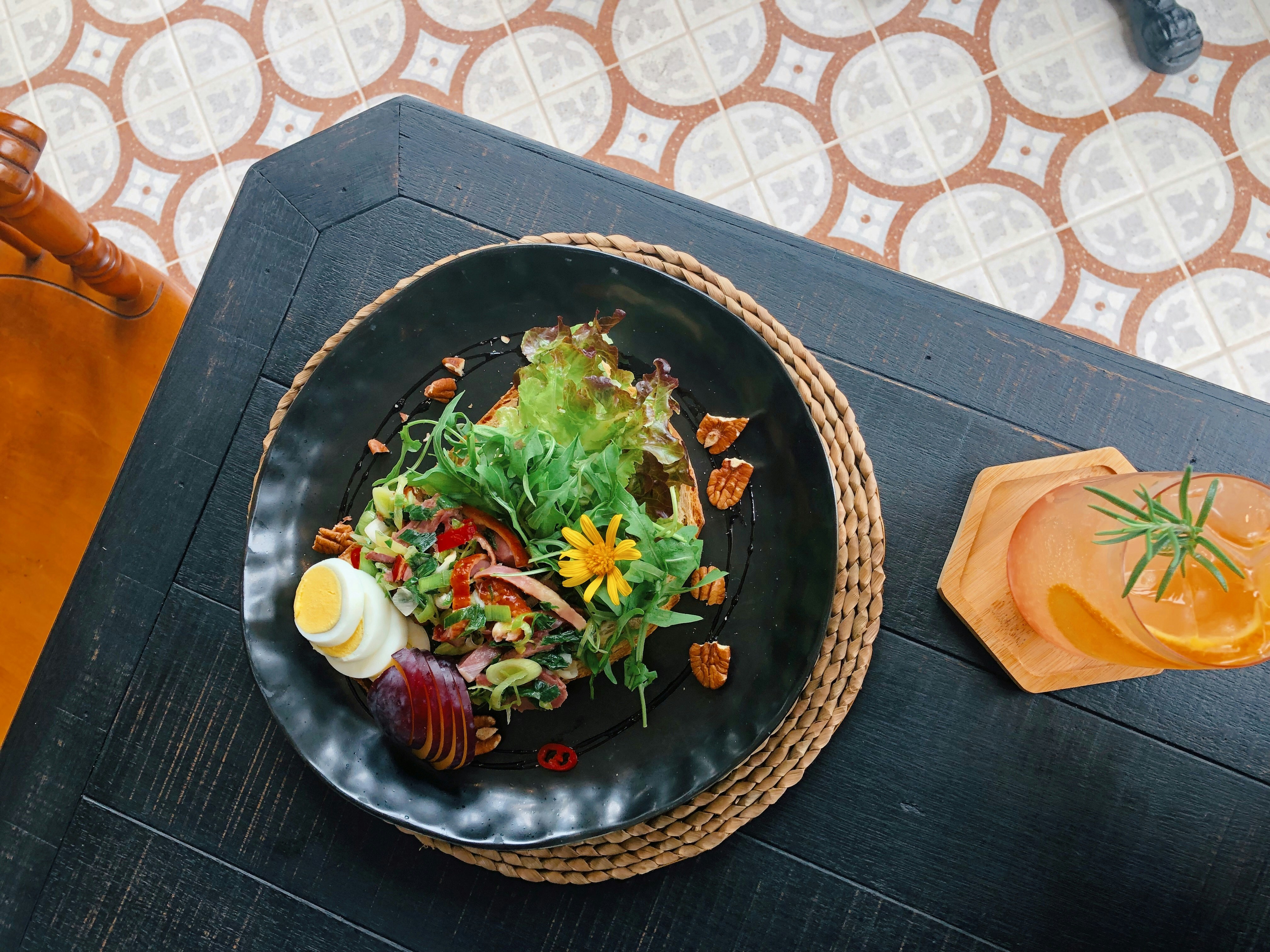 Vibrant salad featuring fresh greens, colorful vegetables, and a perfectly cooked egg, artfully arranged on a black plate. Accompanied by a refreshing drink on the side.