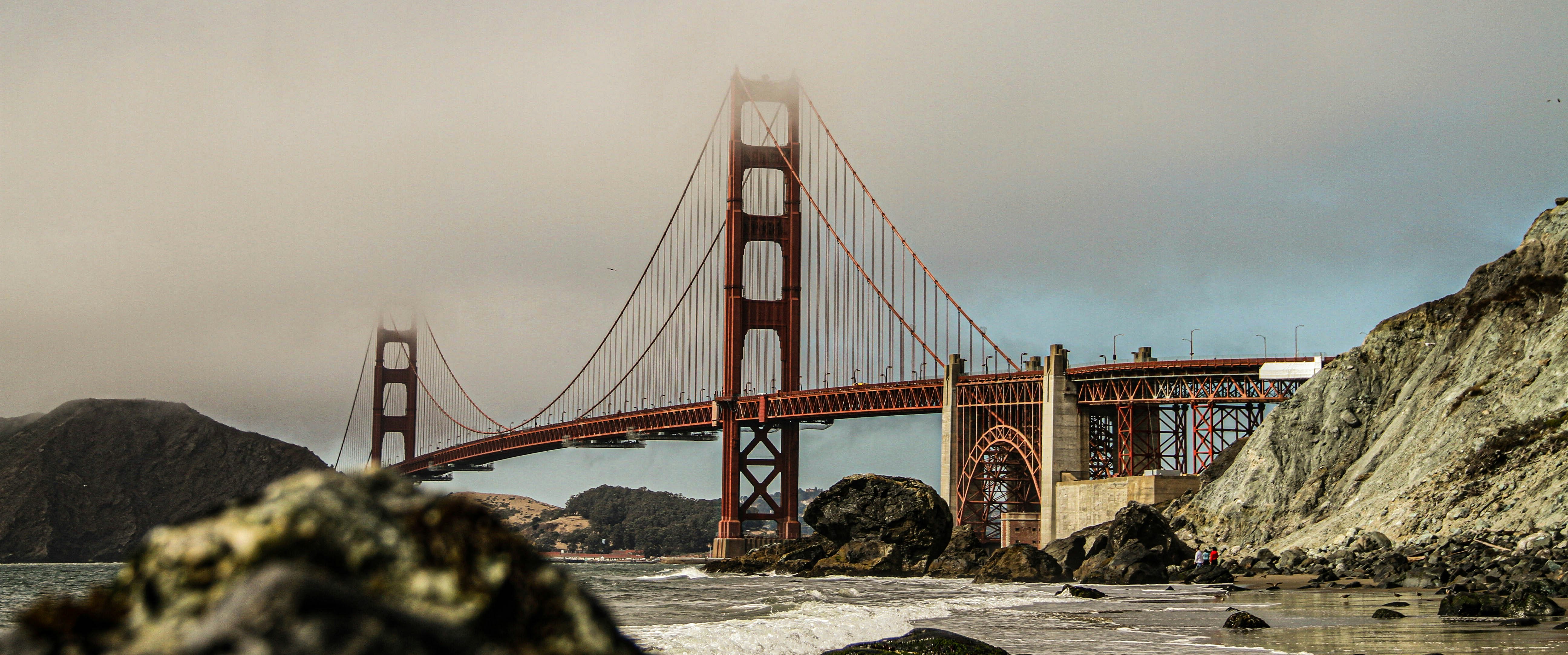 Golden Gate Bridge partially shrouded in fog, framed by rocky shoreline and gentle waves. The iconic structure stands as a testament to engineering and beauty.