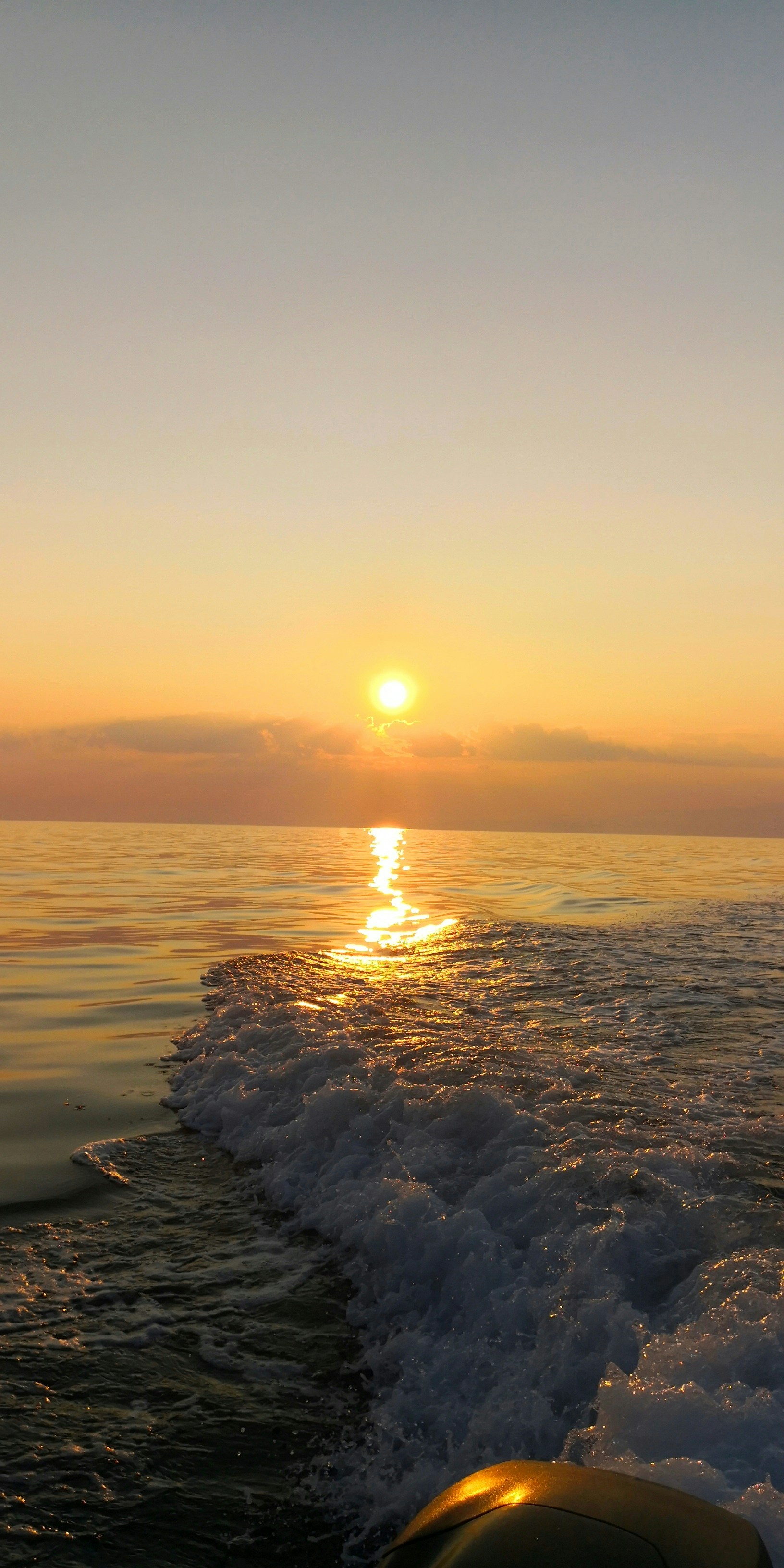Golden sun setting over a calm sea, with gentle waves trailing behind a boat. The horizon glows with warm hues of orange and pink.