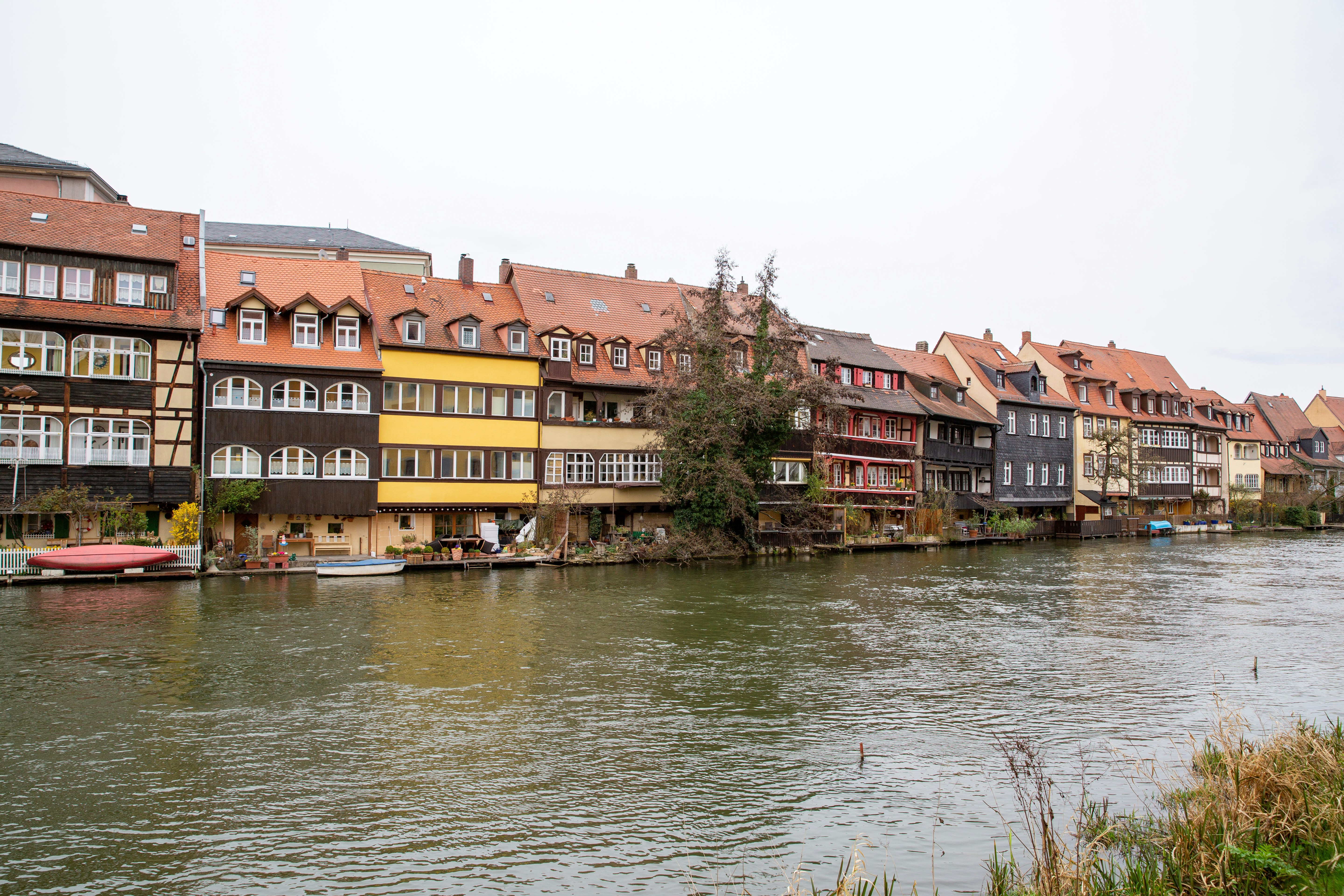 Historic half-timbered houses line a riverside under an overcast sky, reflecting in the gentle flow of the water.