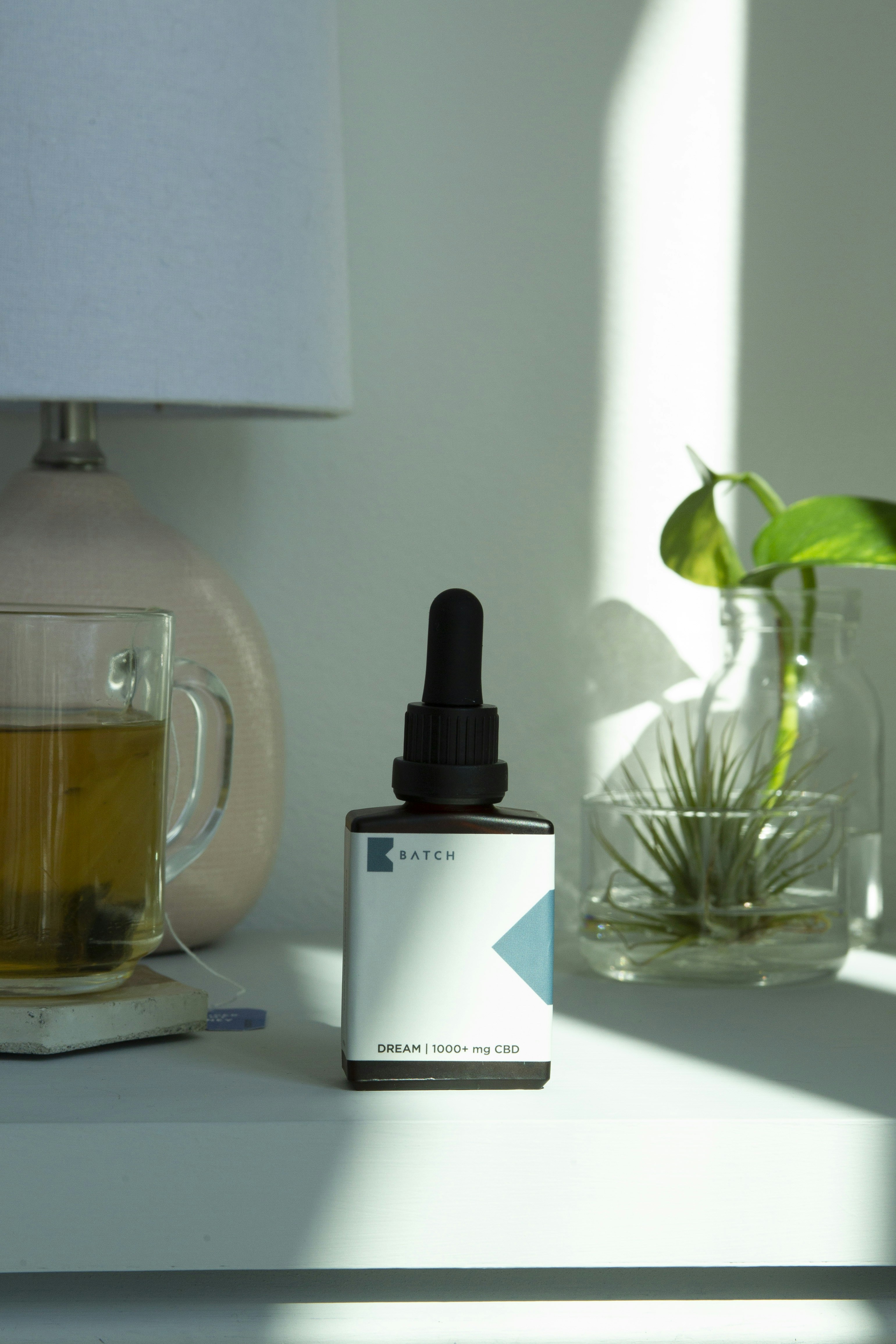 Dark dropper bottle labeled 'BATCH' sits center on a white shelf beside a glass mug of tea and a plant-filled glass vase, bathed in soft daylight. This minimalist still-life composition highlights a product presentation.