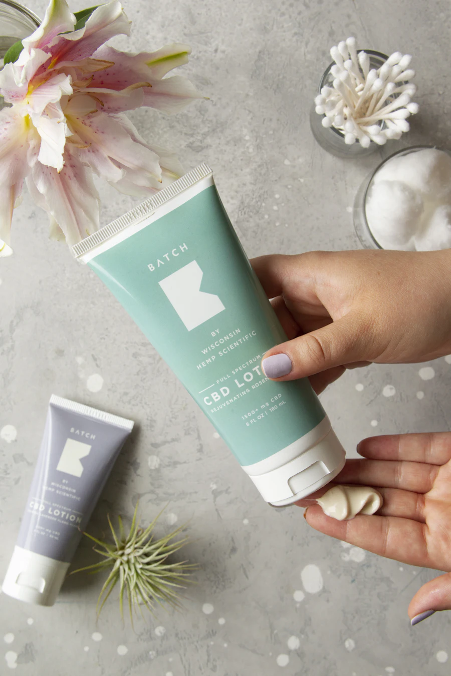 Hands present a turquoise Batch CBD lotion tube with a dollop of cream on the palm below. Soft florals, cotton swabs, and a second lotion tube decorate the light gray surface.