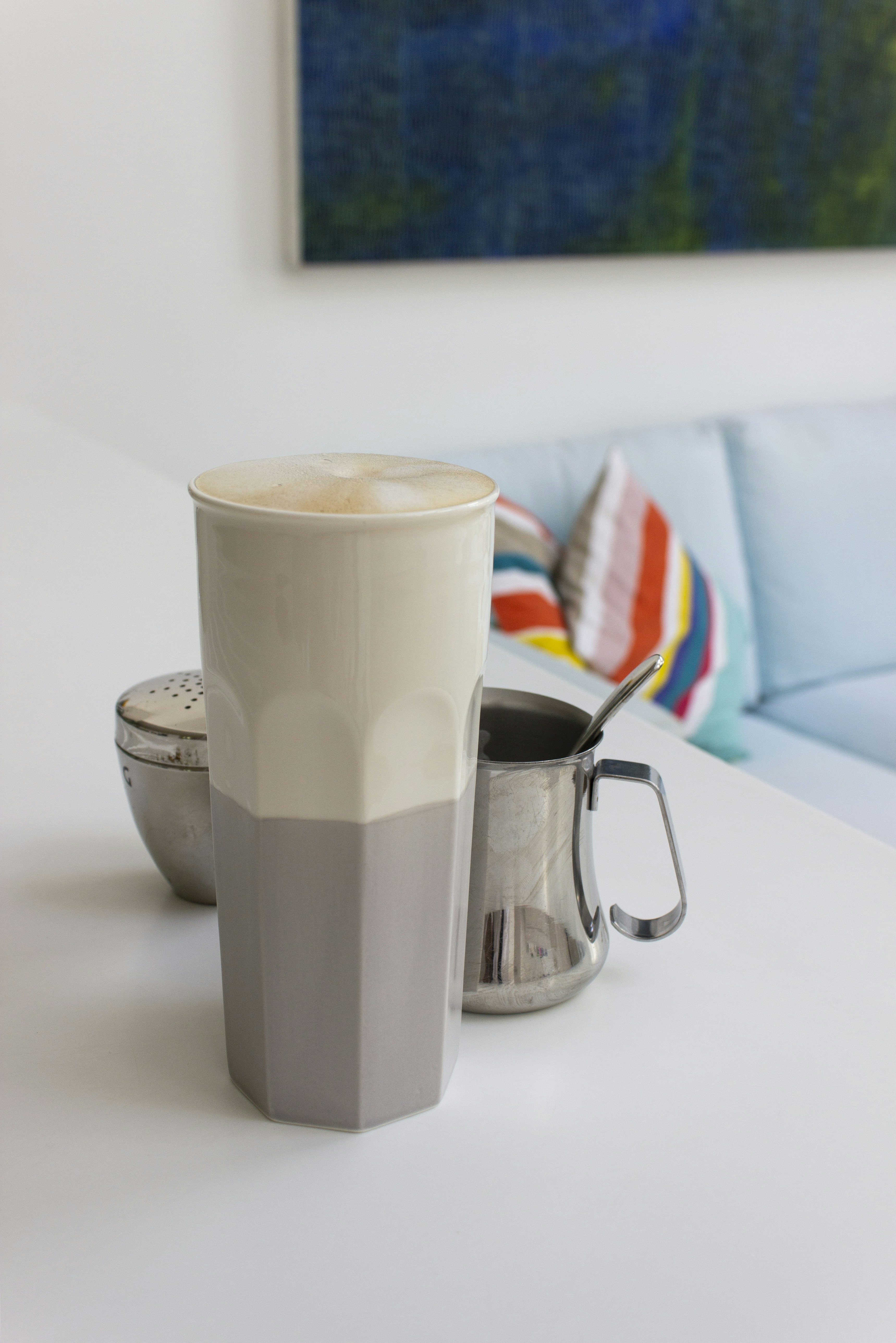 A tall, stylish coffee cup rests on a sleek table, accompanied by a milk frother and a sugar container, with a cozy couch and colorful cushions in the background.