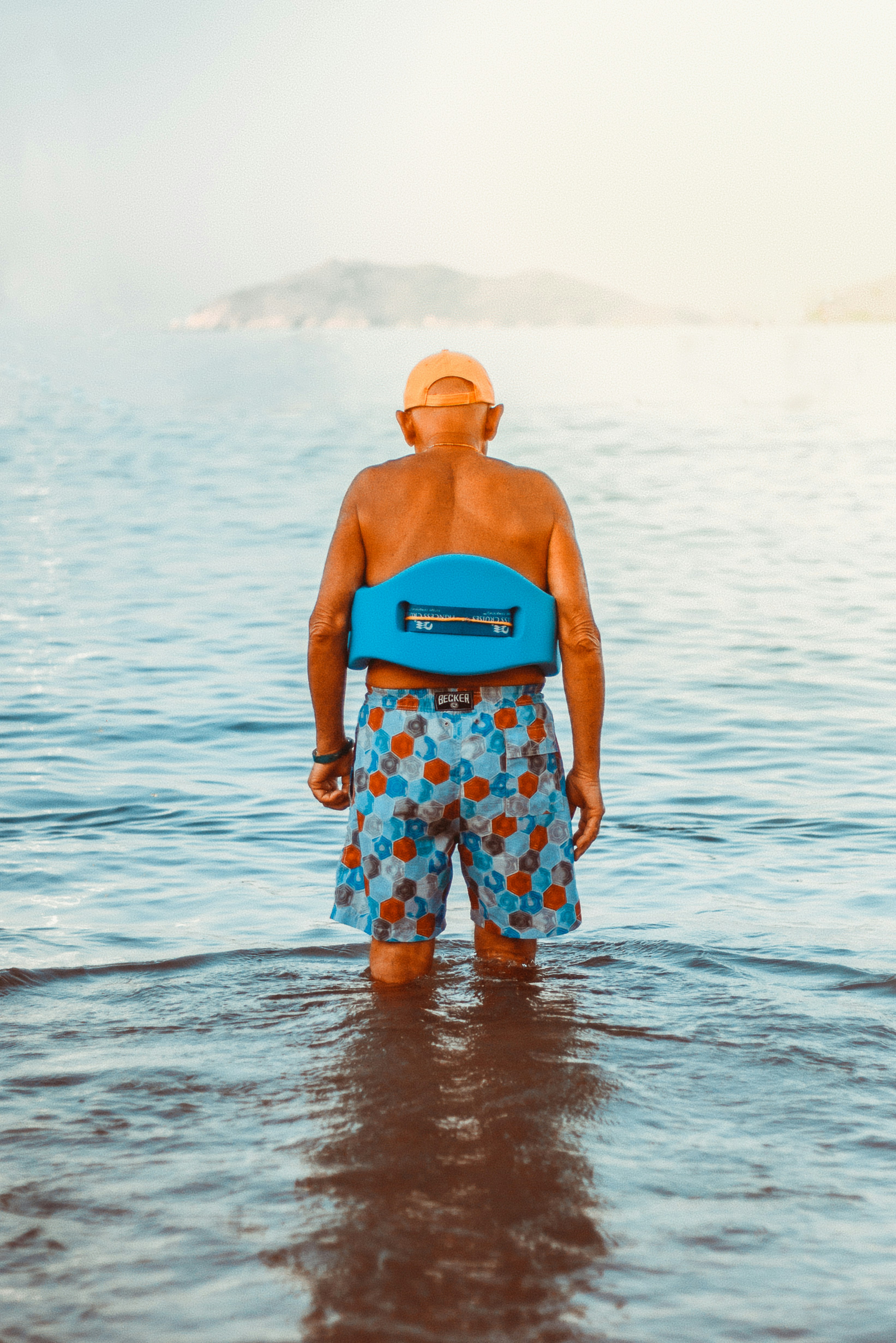 Elderly man wading into calm waters, wearing colorful swim shorts and a buoyancy aid, with distant hills fading into the horizon.
