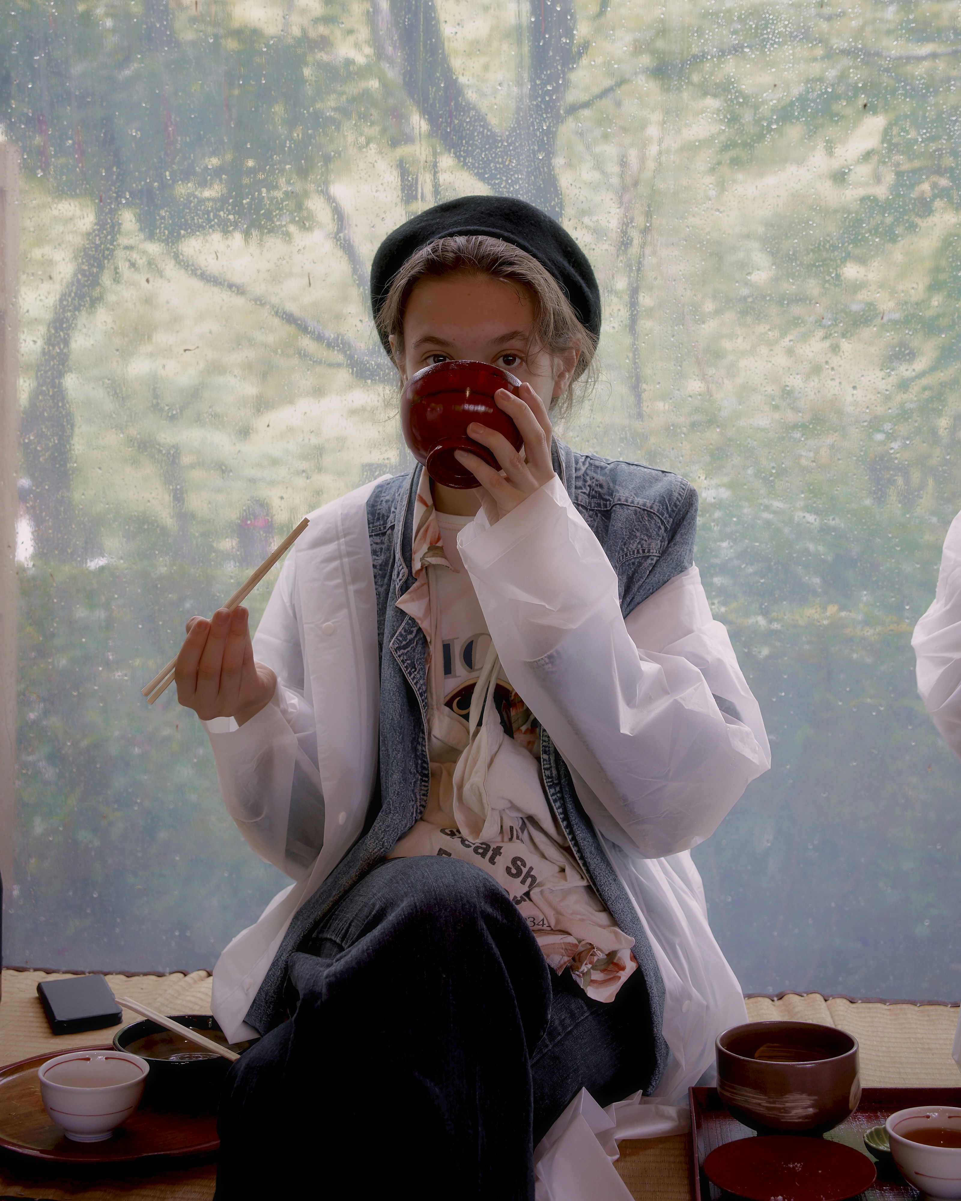 Young woman enjoying a bowl of food while seated at a table, with a serene outdoor backdrop blurred by raindrops on glass.