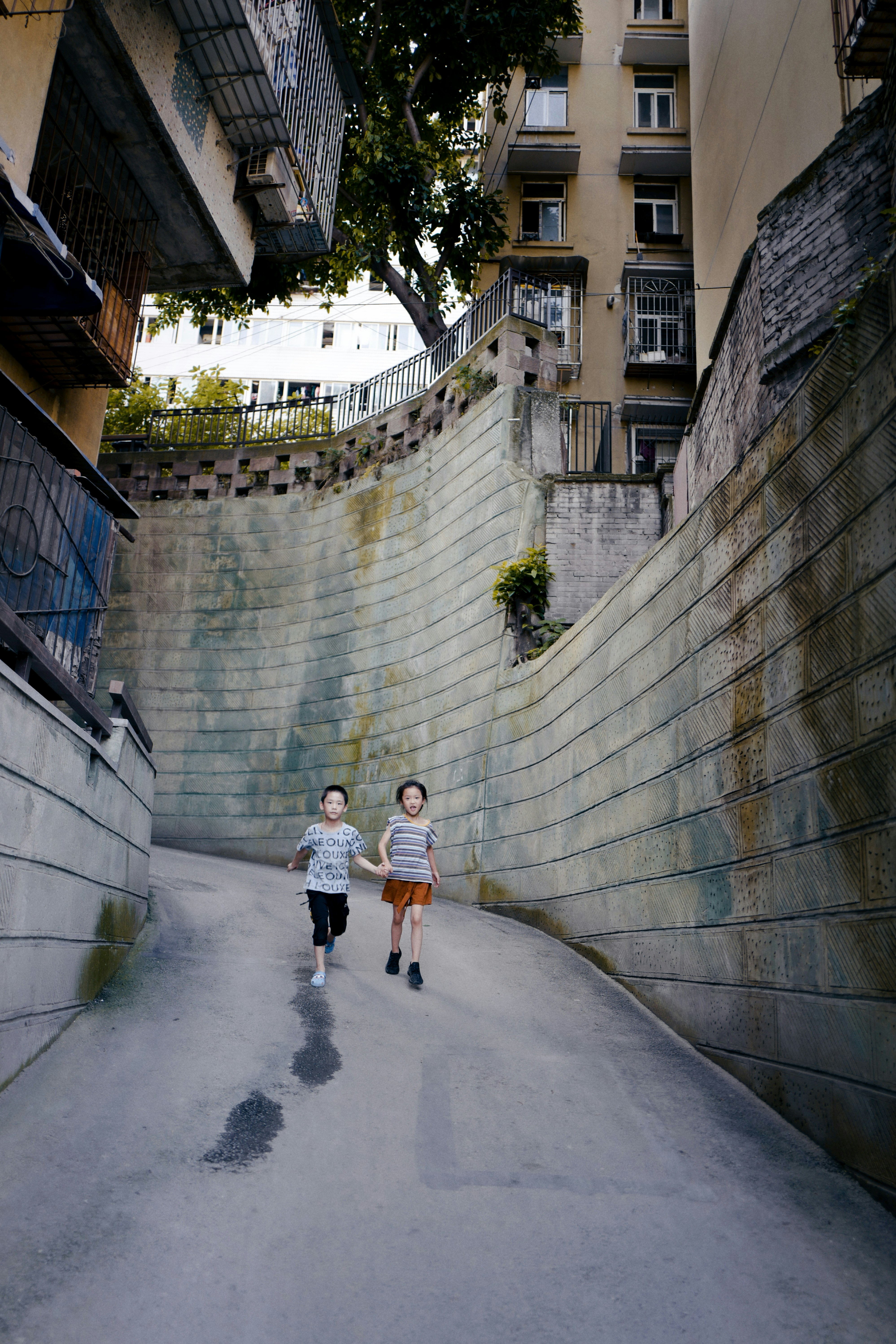 Two children joyfully running down a winding urban path, flanked by textured concrete walls and greenery above.