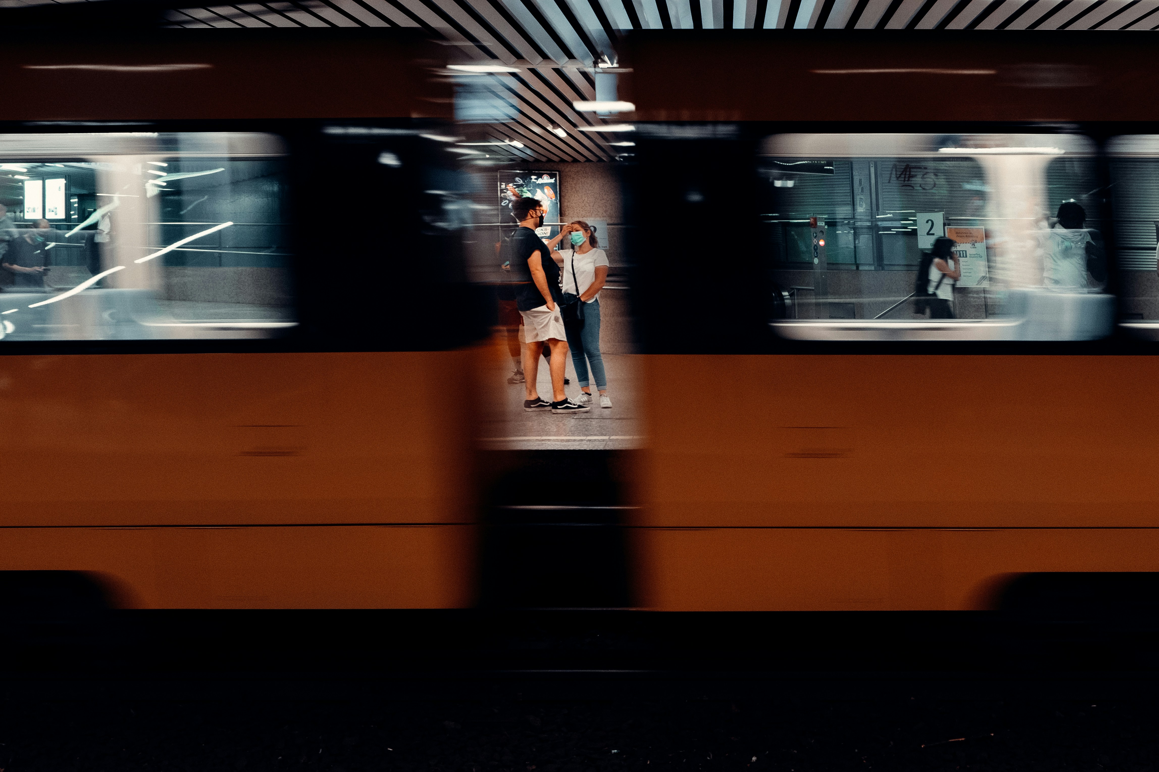 A couple stands on the platform as a train rushes past, capturing the fleeting nature of urban life. The motion blur emphasizes the speed of the train contrasted with the stillness of the moment.