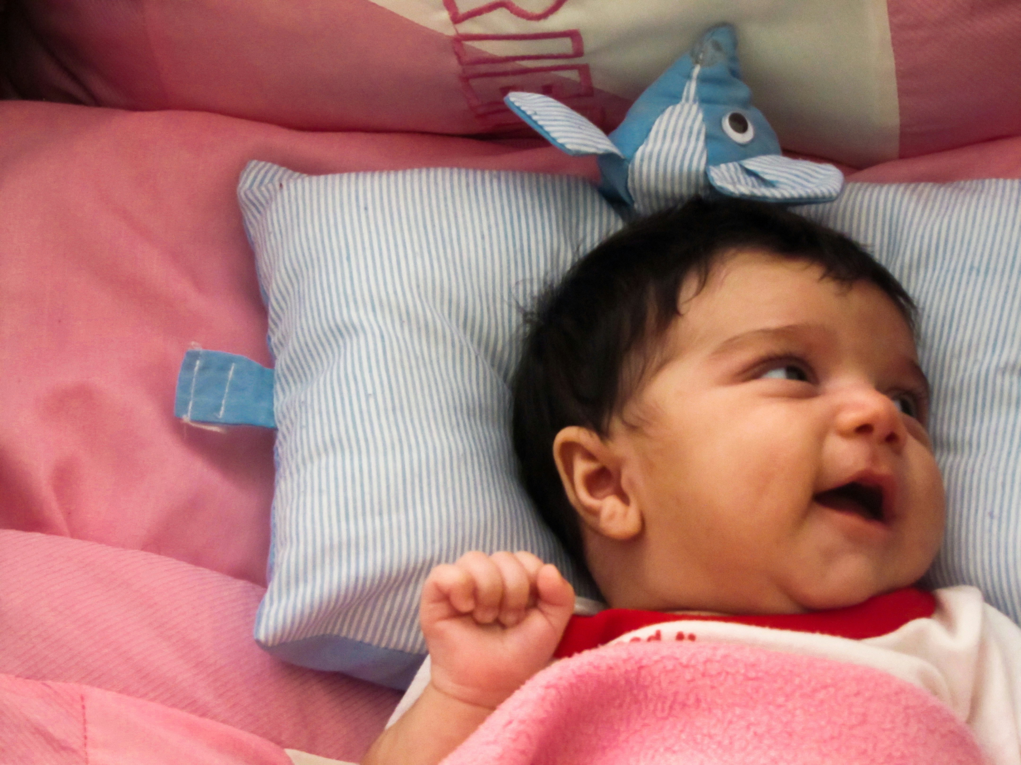 Infant lying on pink bedding, smiling while holding a small fist, with a plush fish toy above. 